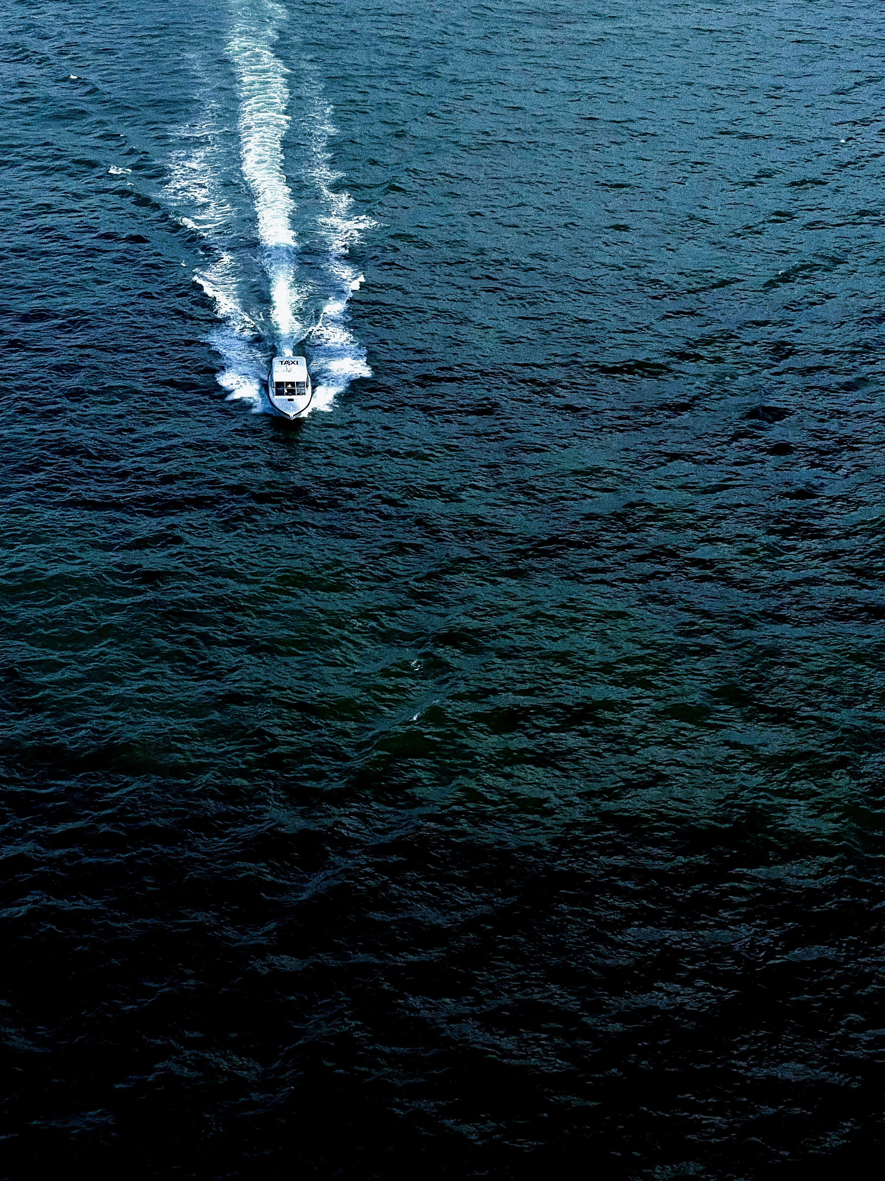 A boat glides across a deep blue sea, leaving a trail of white foam in its wake. The serene water surface reflects varying shades of blue.