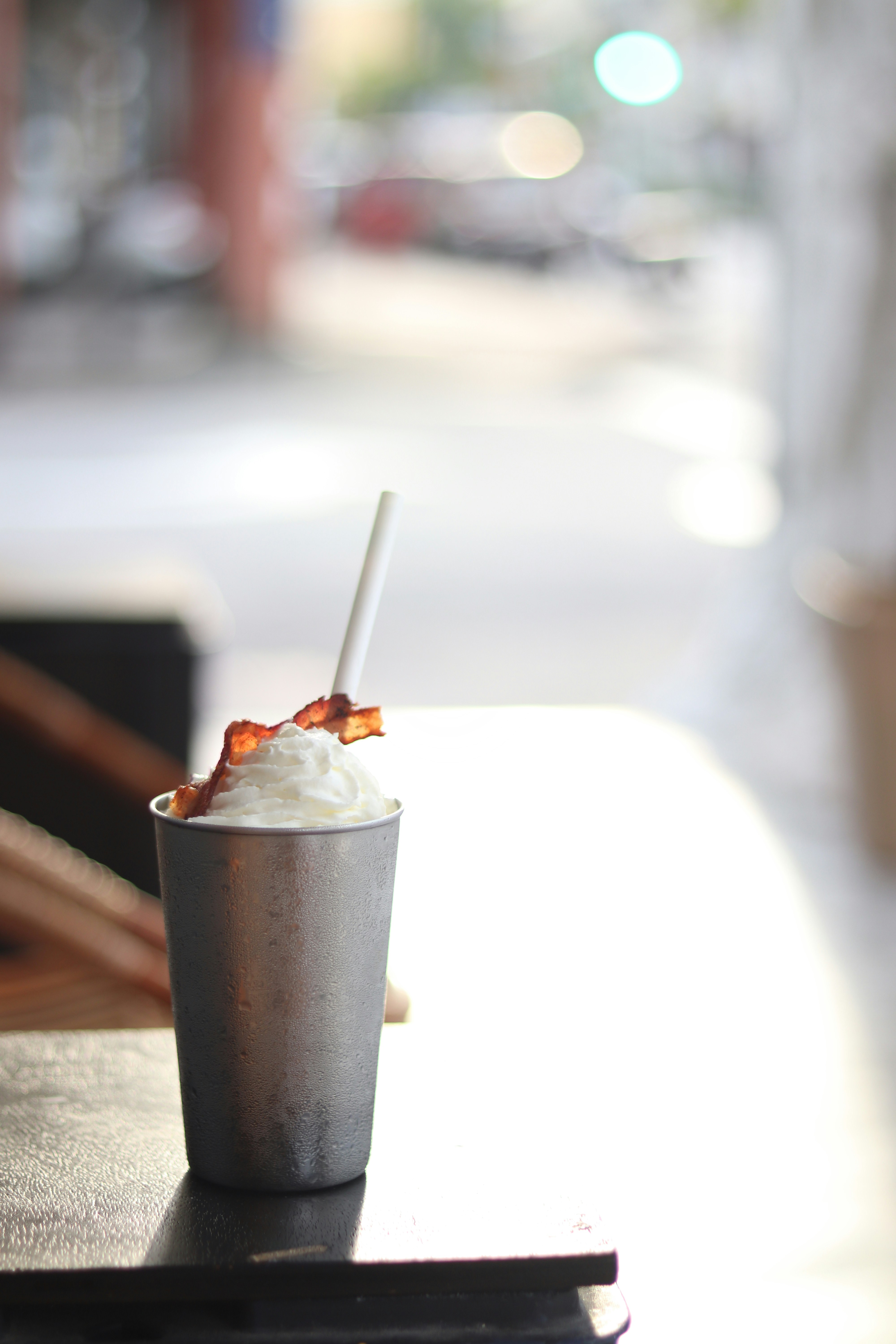 Metal cup filled with whipped cream and crispy bacon, set against a softly blurred urban background. 