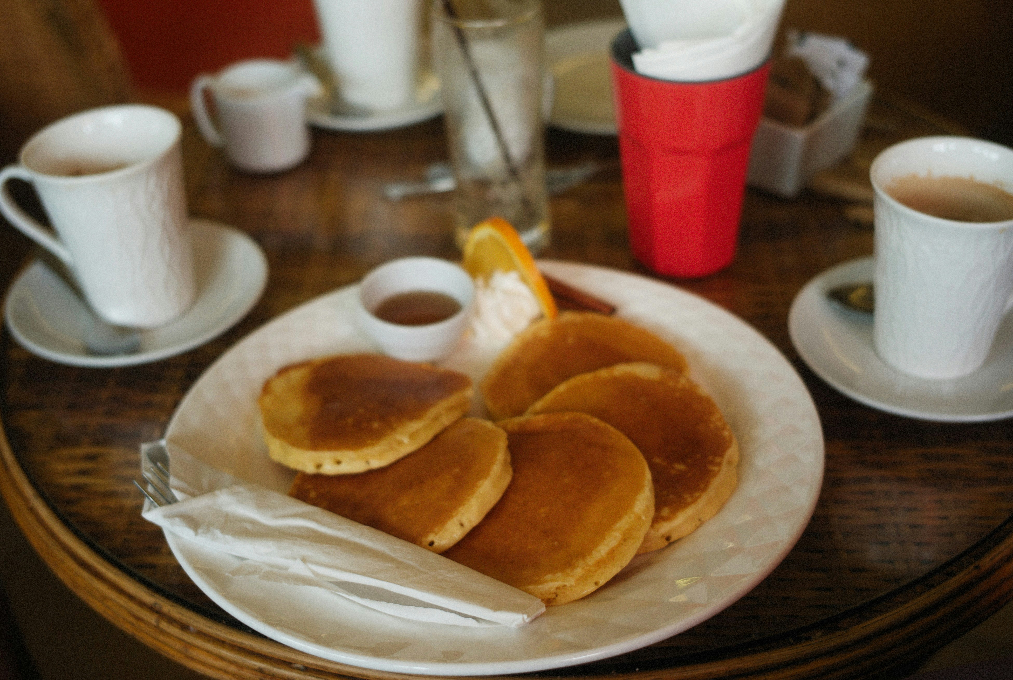 A breakfast scene on a wooden table featuring a plate of fluffy pancakes with syrup and a lemon wedge. Several cups of coffee and a red napkin holder frame the composition.