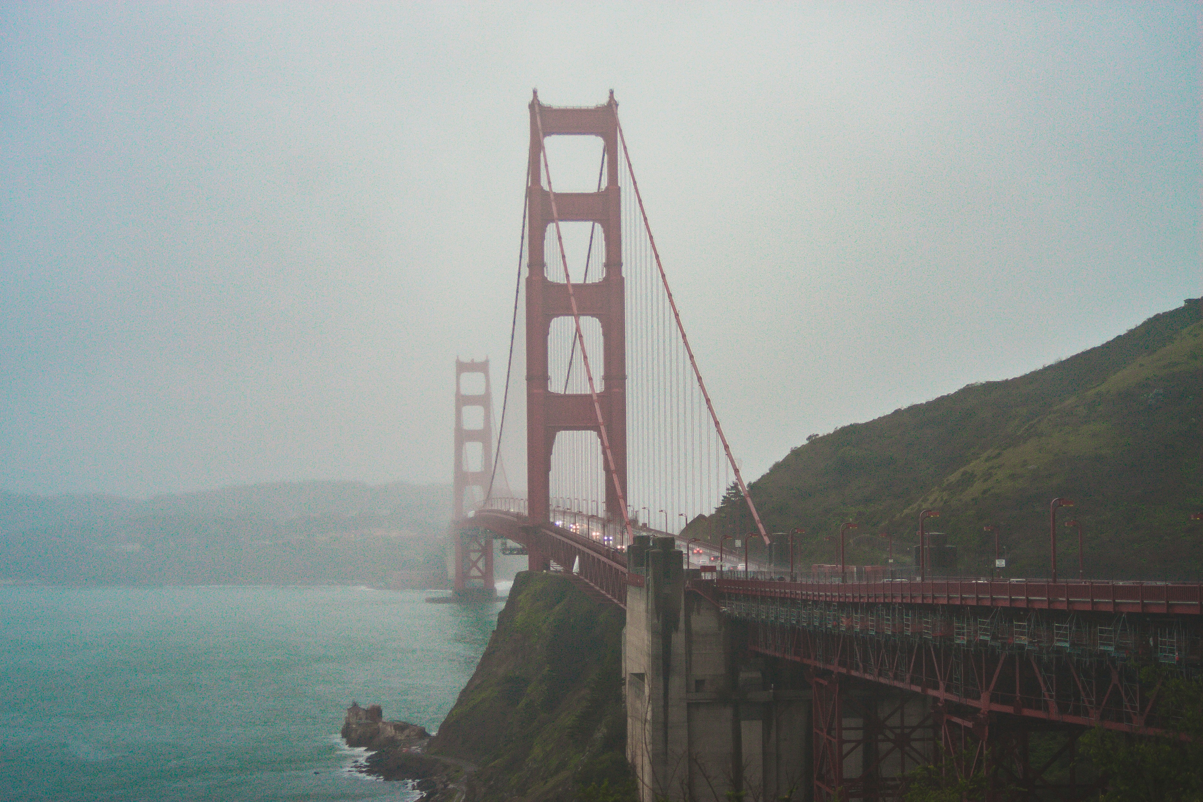 Golden Gate Bridge shrouded in fog, with lush green hills framing the scene and waves crashing against the rocky shore.