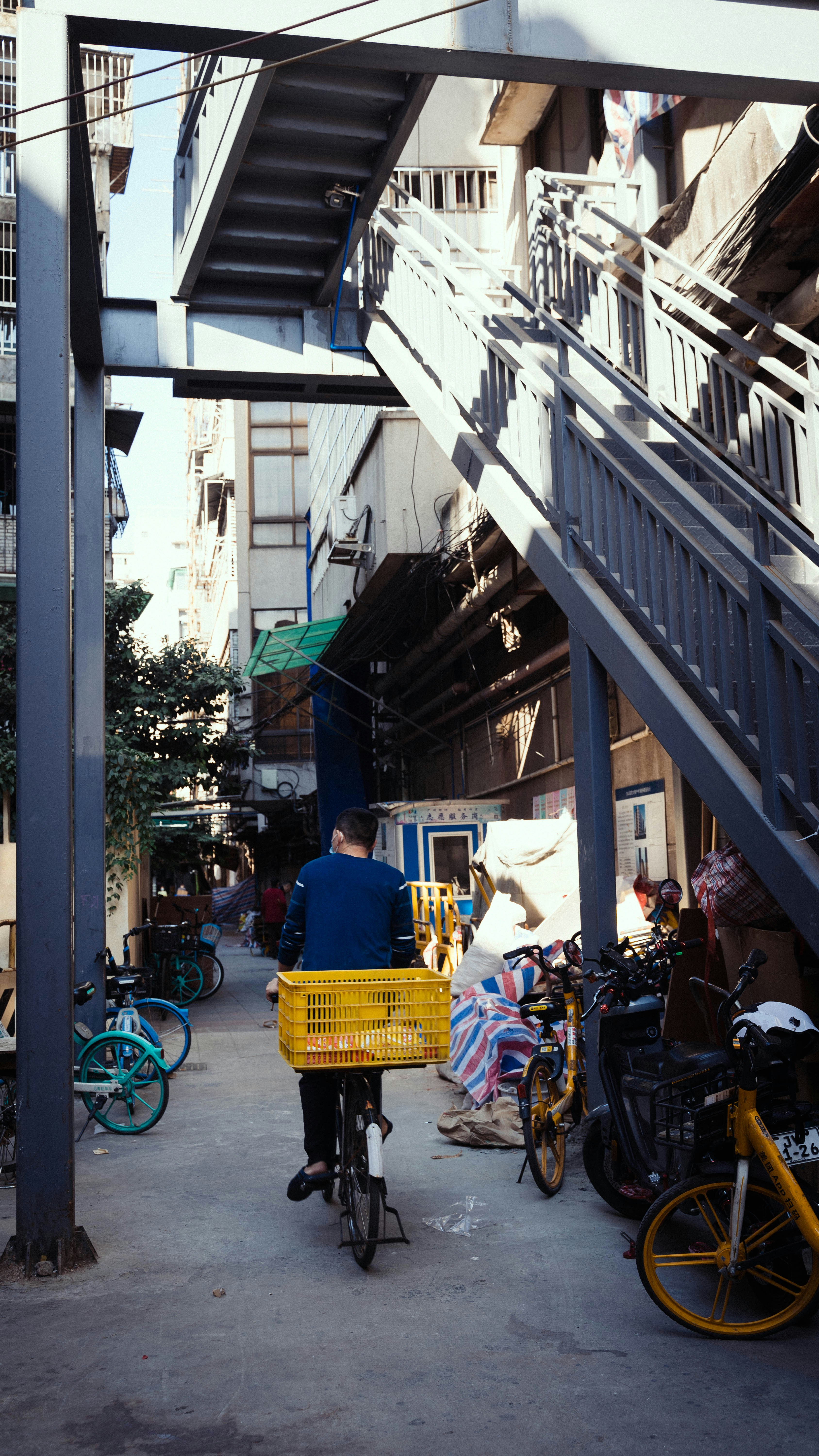 Man cycling through a narrow alleyway adorned with bicycles, framed by a staircase and urban structures. A blend of everyday life and city architecture.