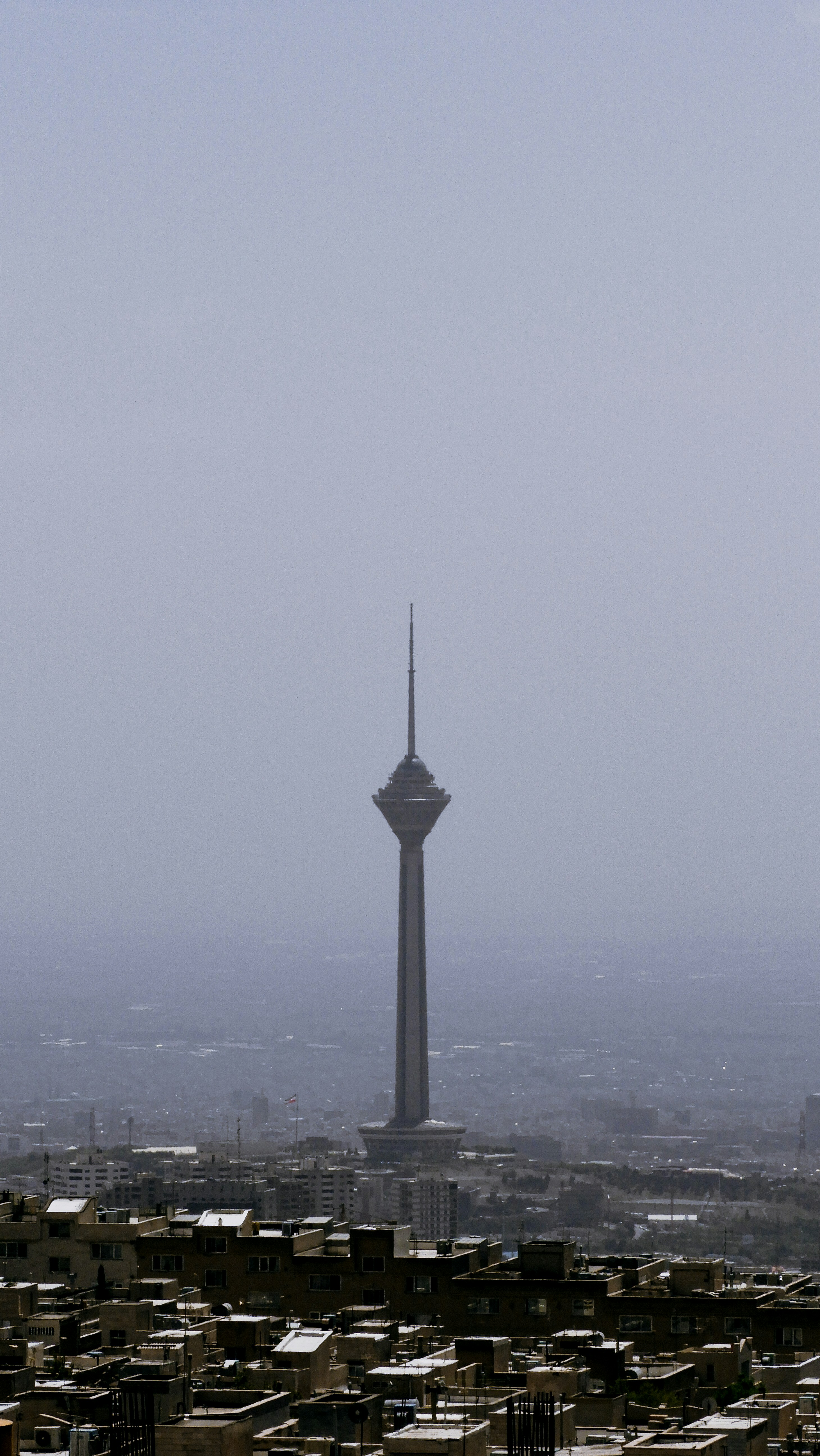 Milad Tower rises above the cityscape of Tehran, surrounded by an expanse of urban structures under a hazy sky.