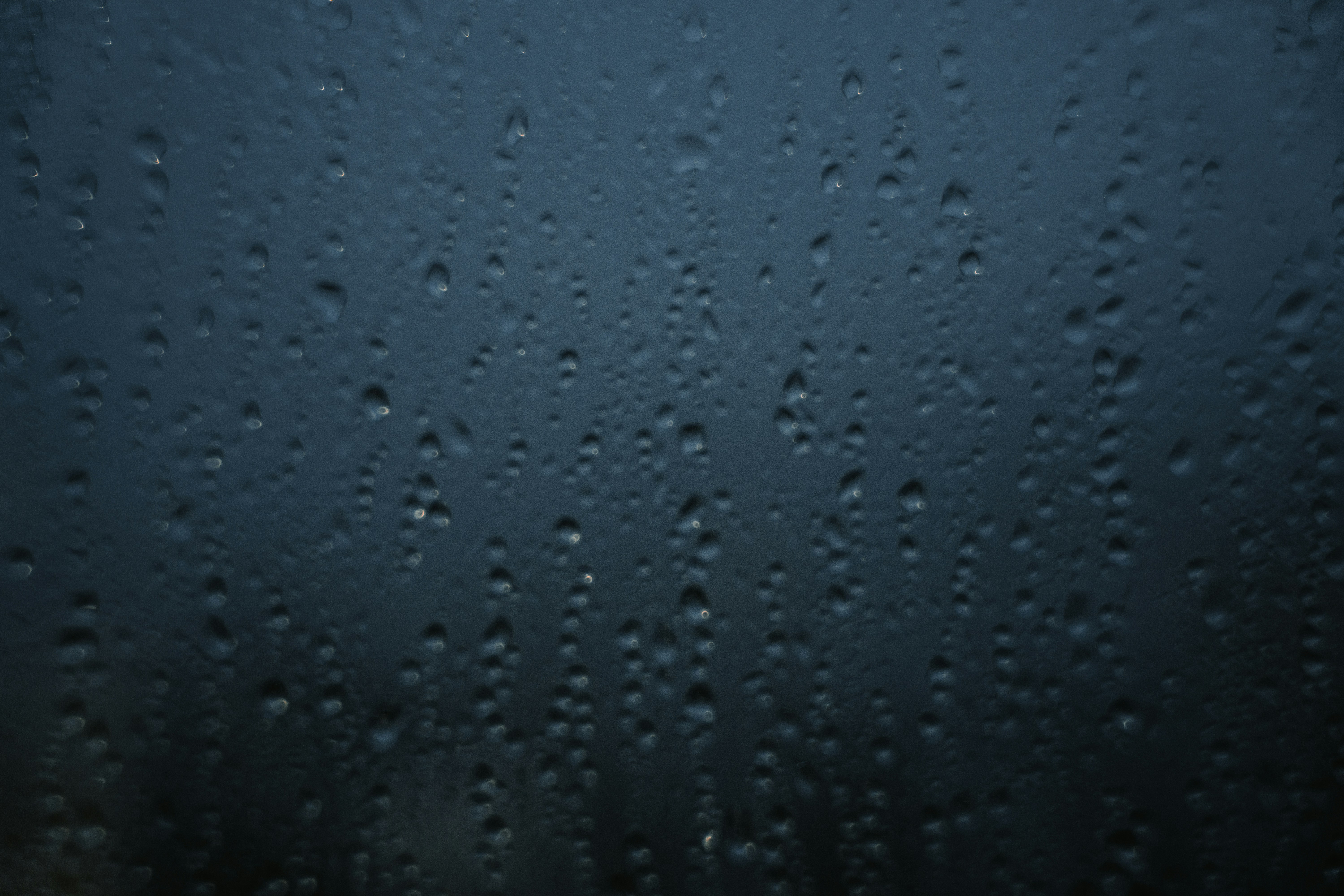 Close-up of raindrops clinging to a glass surface, creating a blurred view of the outside world. The dark background enhances the mood of a rainy day.