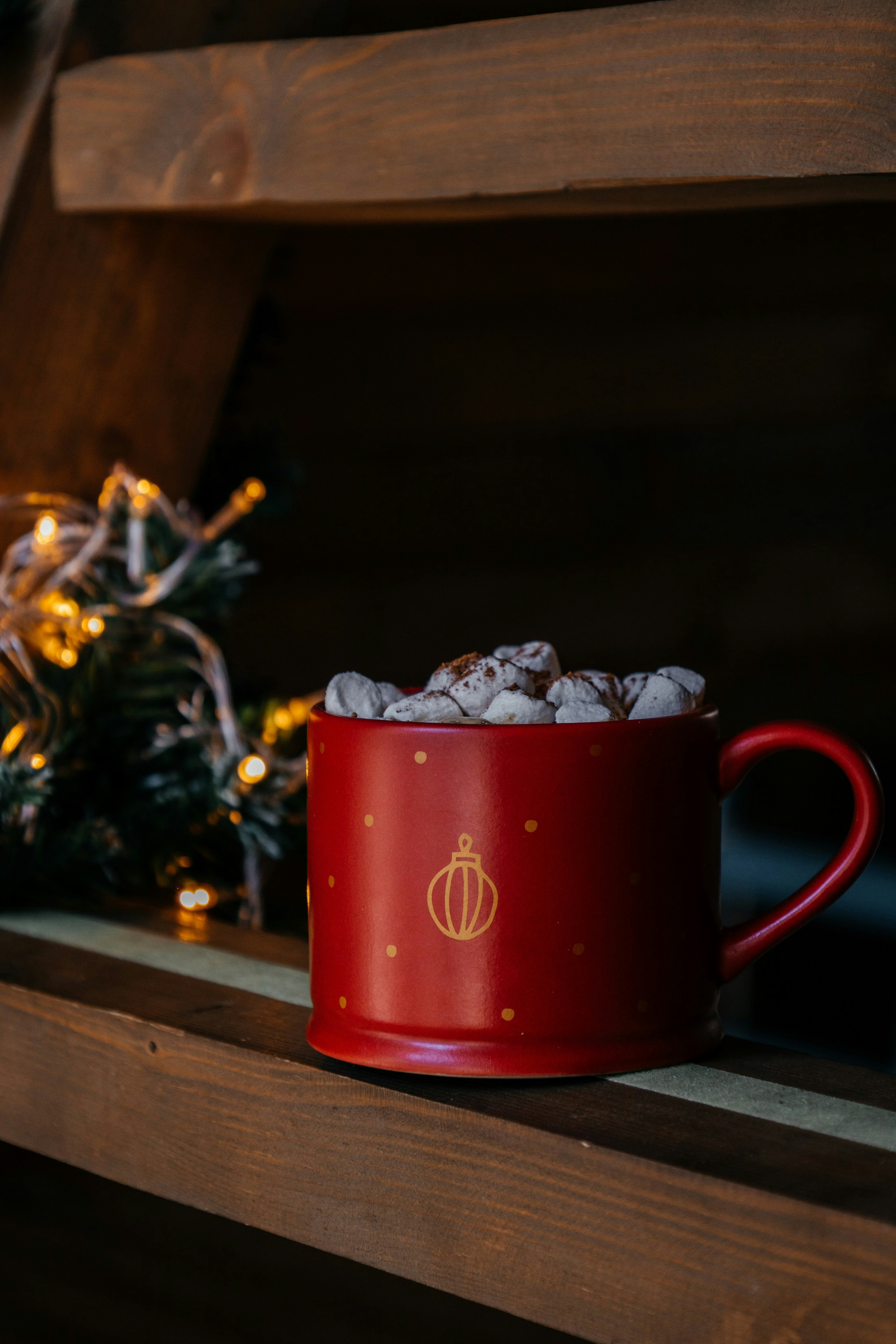Red mug filled with marshmallows sits on a wooden shelf, surrounded by twinkling lights and greenery.