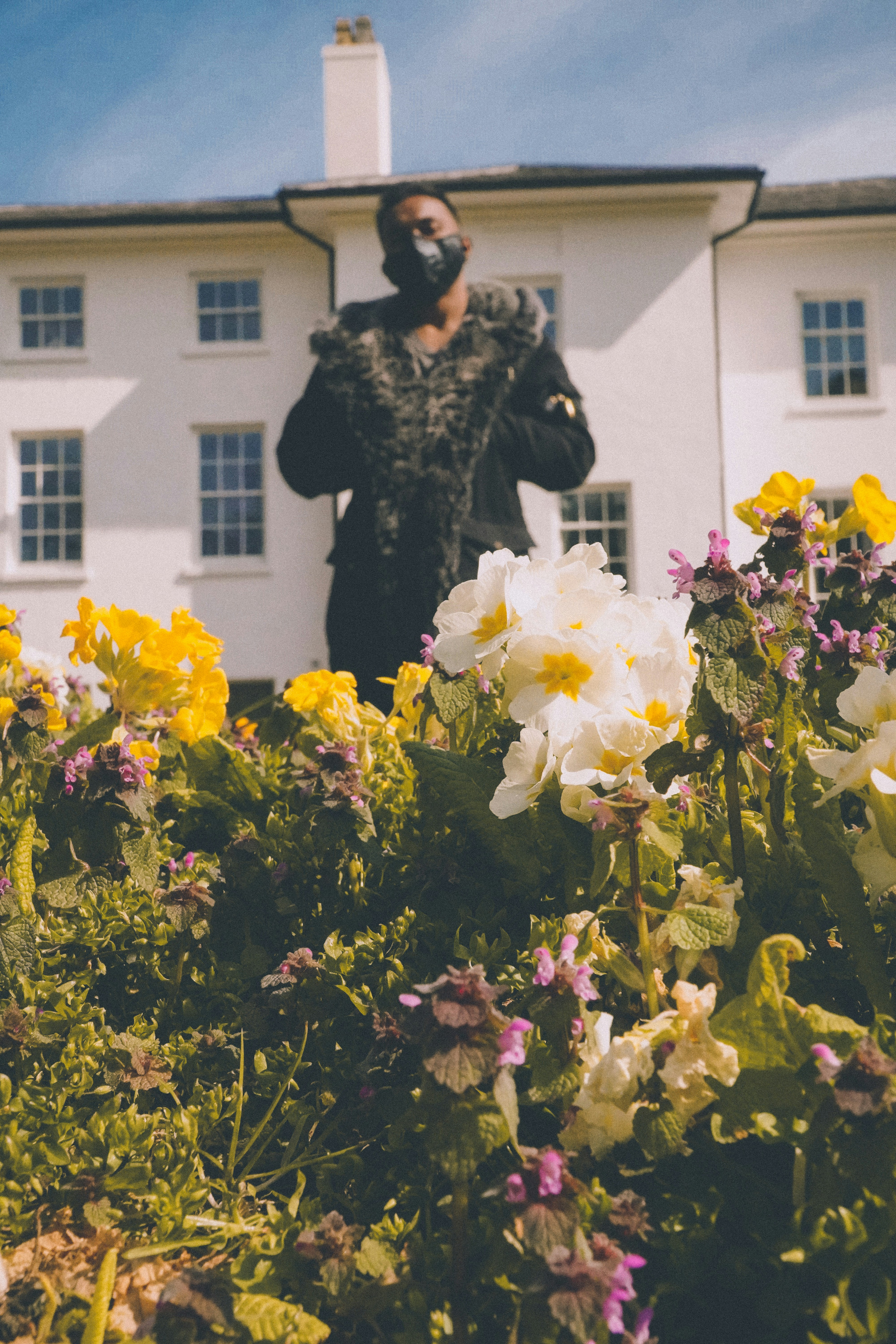 Person in a black coat stands amidst vibrant flowers in a garden, with a white building in the background. The scene captures a blend of nature and urban life.