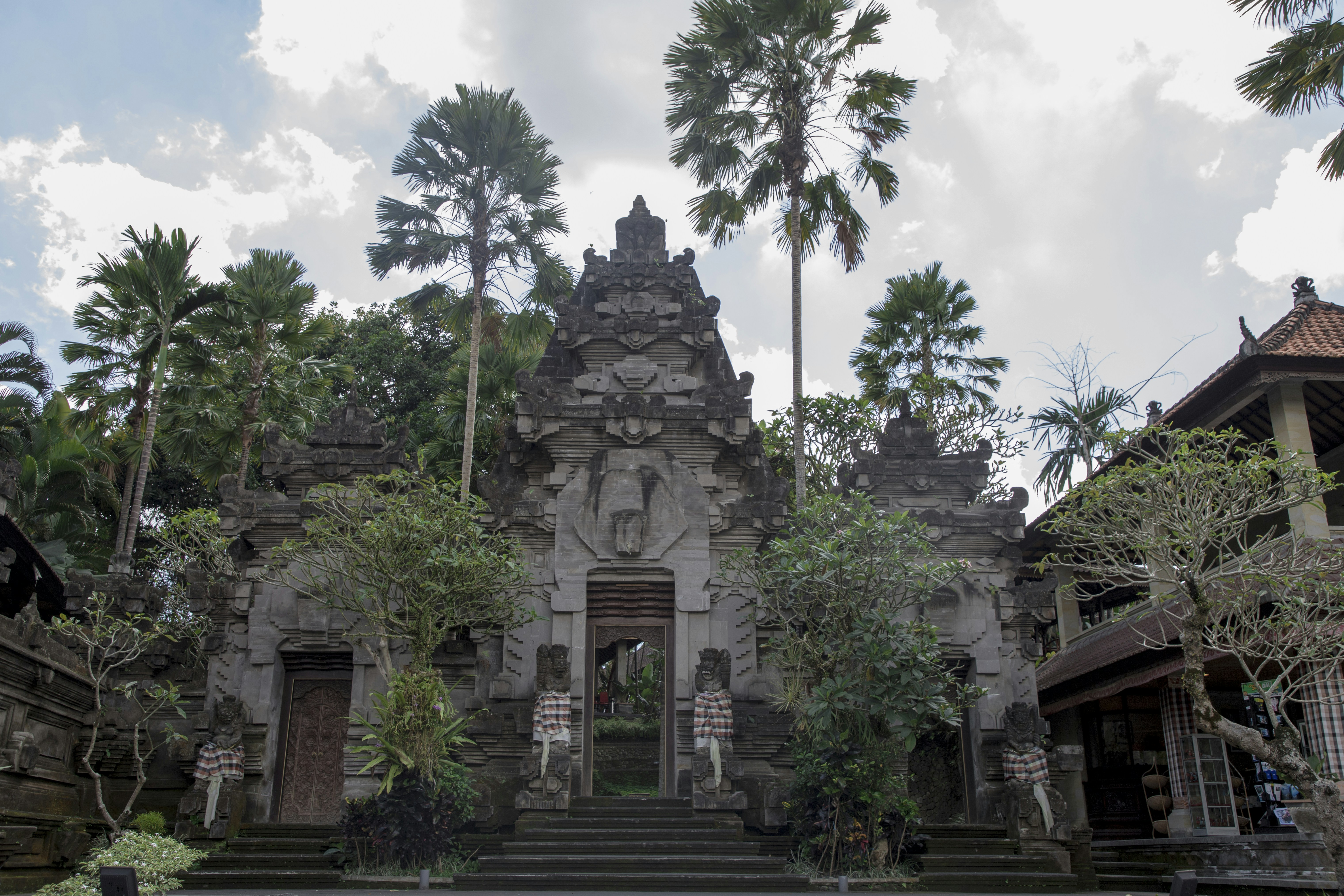 Intricate stone carvings of a Balinese temple framed by lush greenery and palm trees under a cloudy sky.