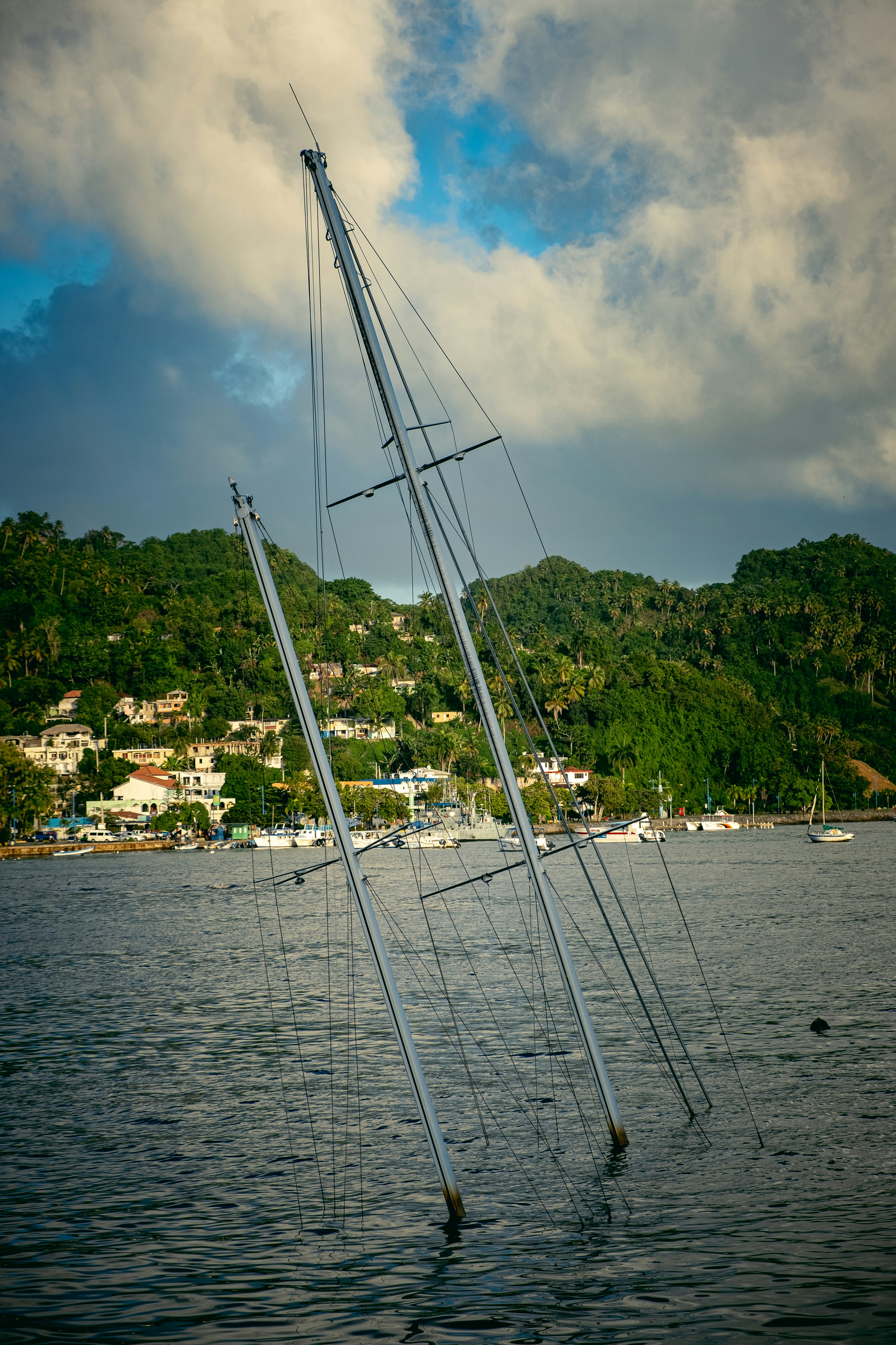 Leaning sailboat masts partially submerged in tranquil waters, framed by lush green hills and coastal homes. 