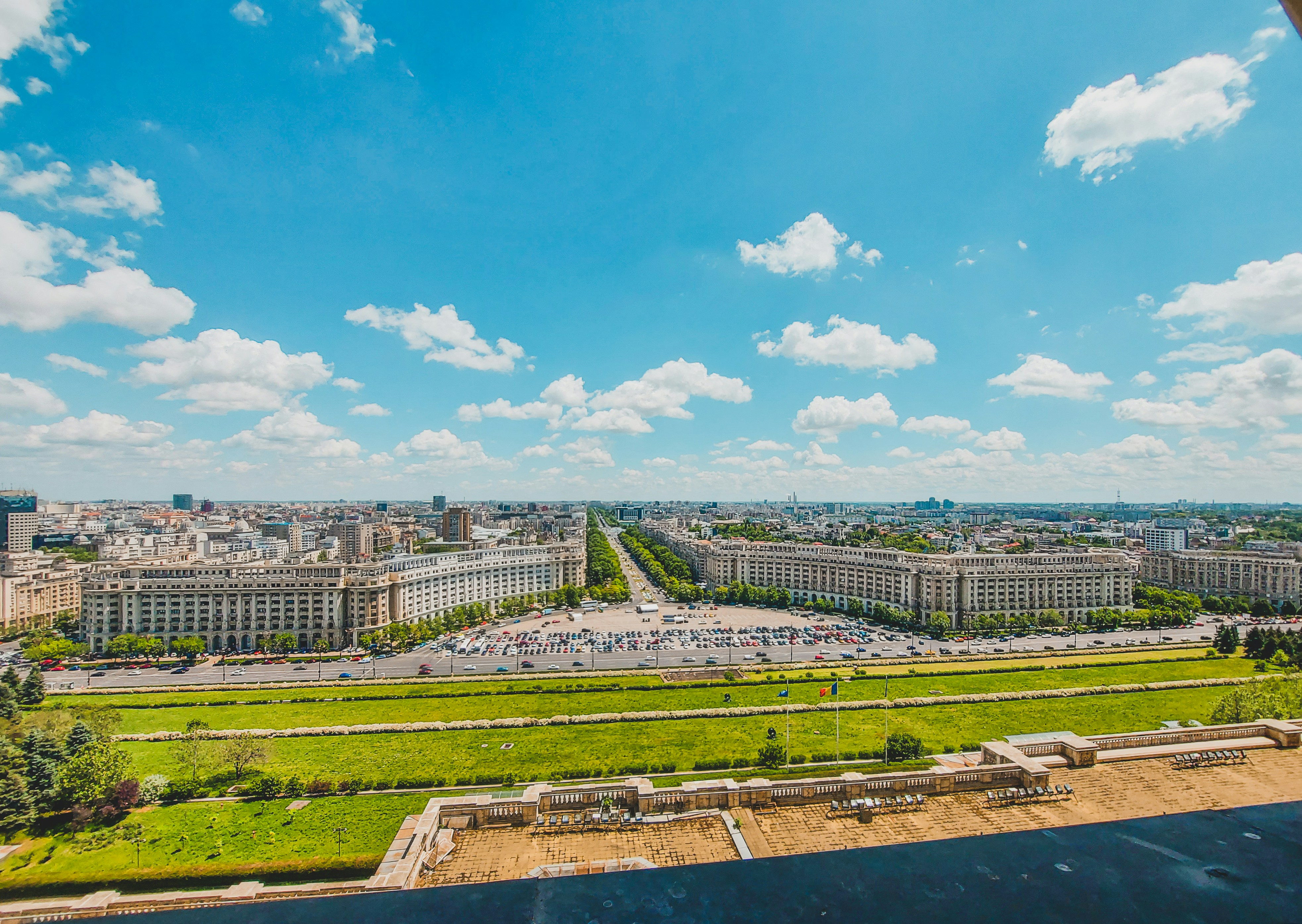 Vast cityscape featuring a wide avenue lined with greenery and buildings under a bright blue sky with fluffy clouds.