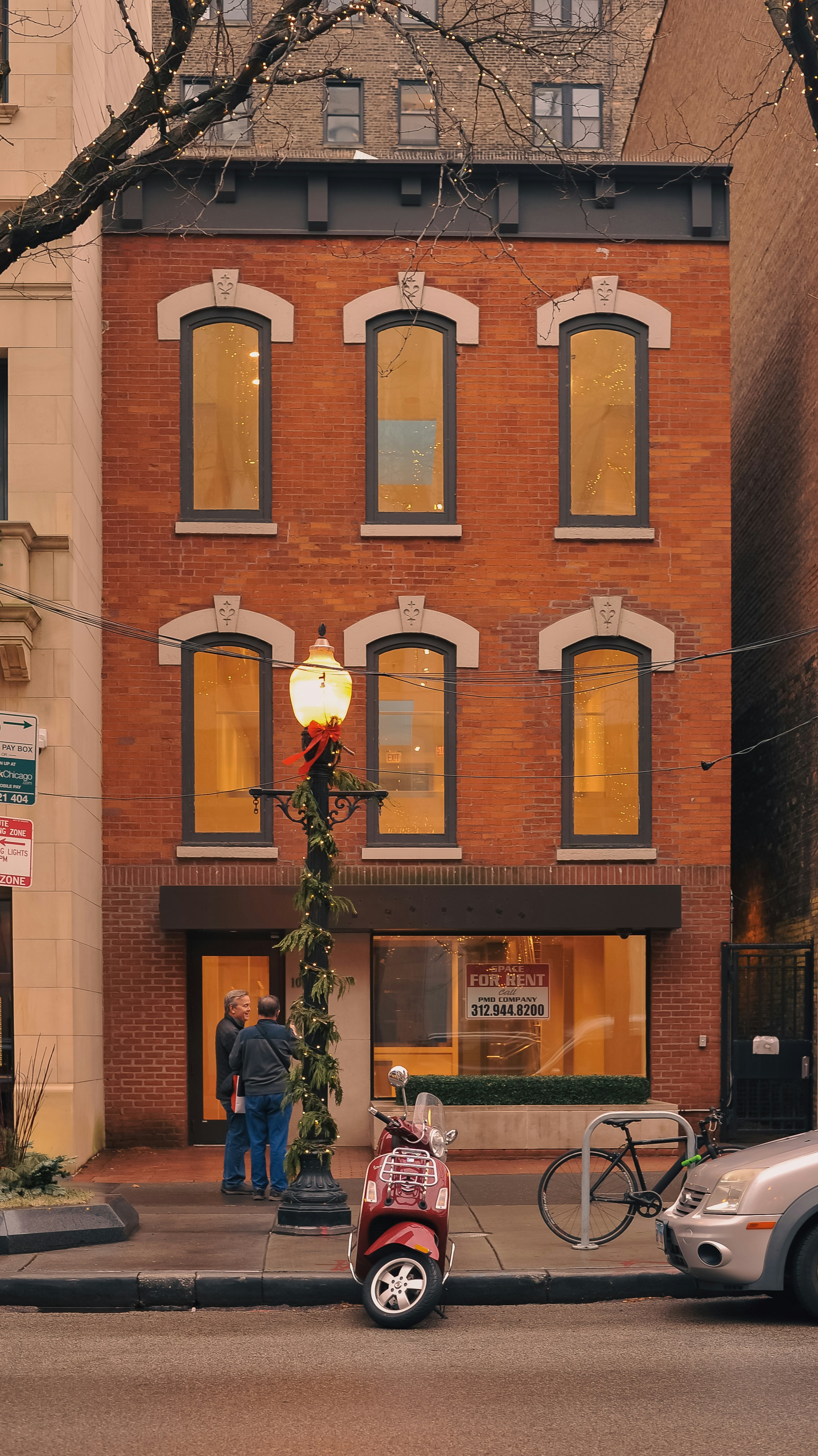 Two individuals converse outside a brick building adorned with holiday decor, while a scooter and bicycle are parked nearby.