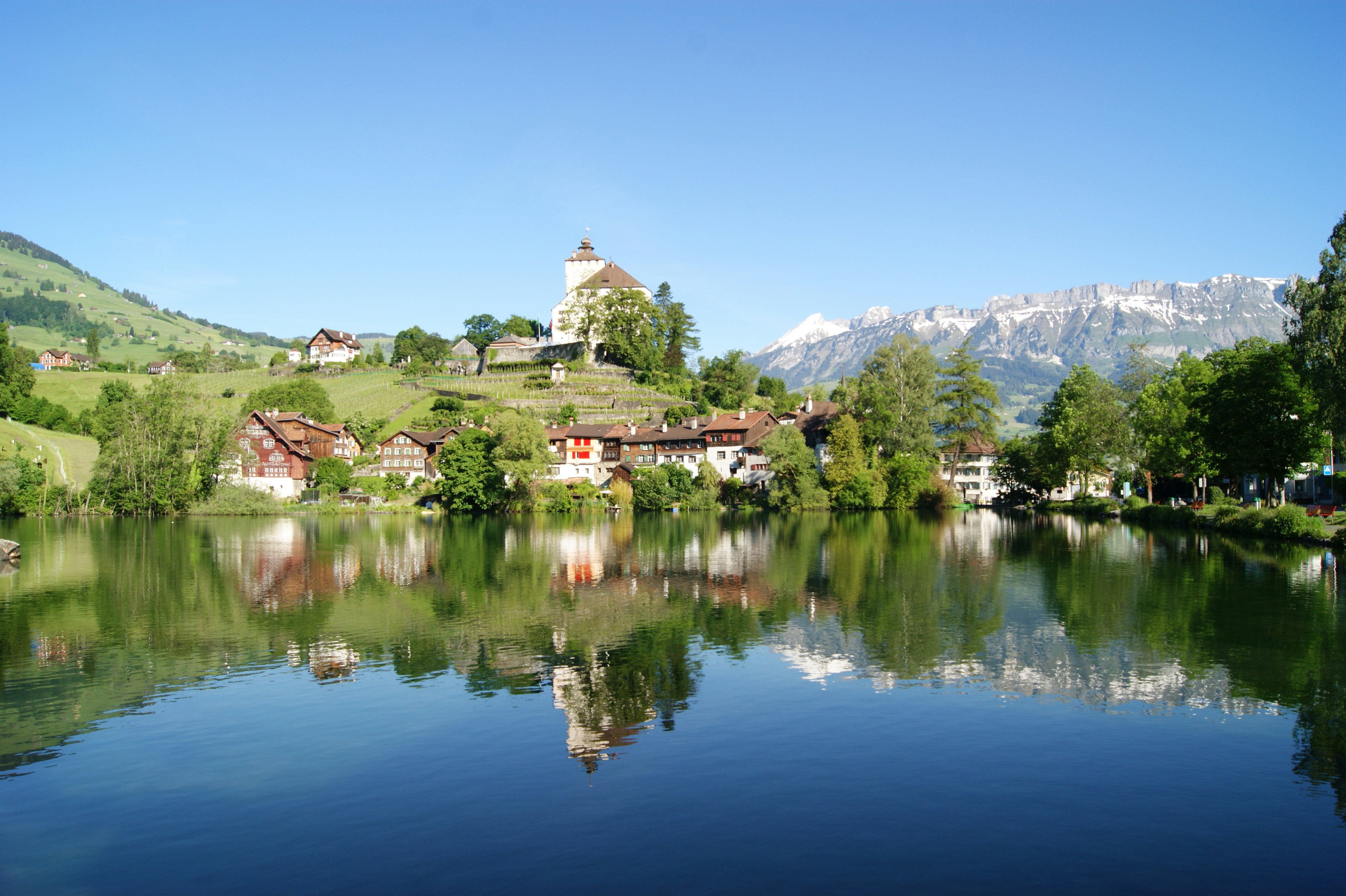 Serene lake reflecting the village of Werdenberg, historic castle, and distant snow-capped mountains under a clear blue sky.