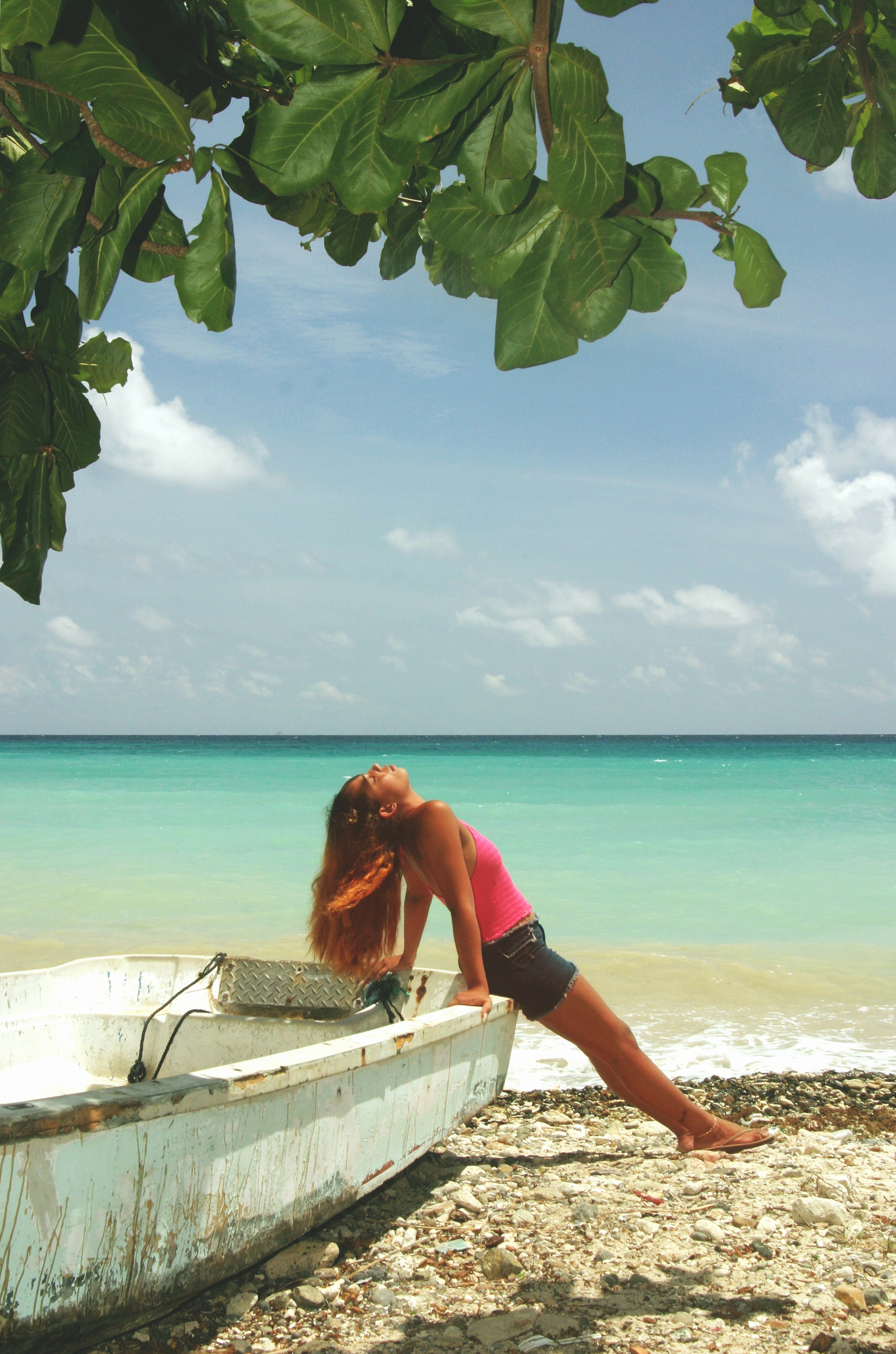 A woman leans back against a weathered boat on a rocky beach, with turquoise waters and a clear sky in the background.