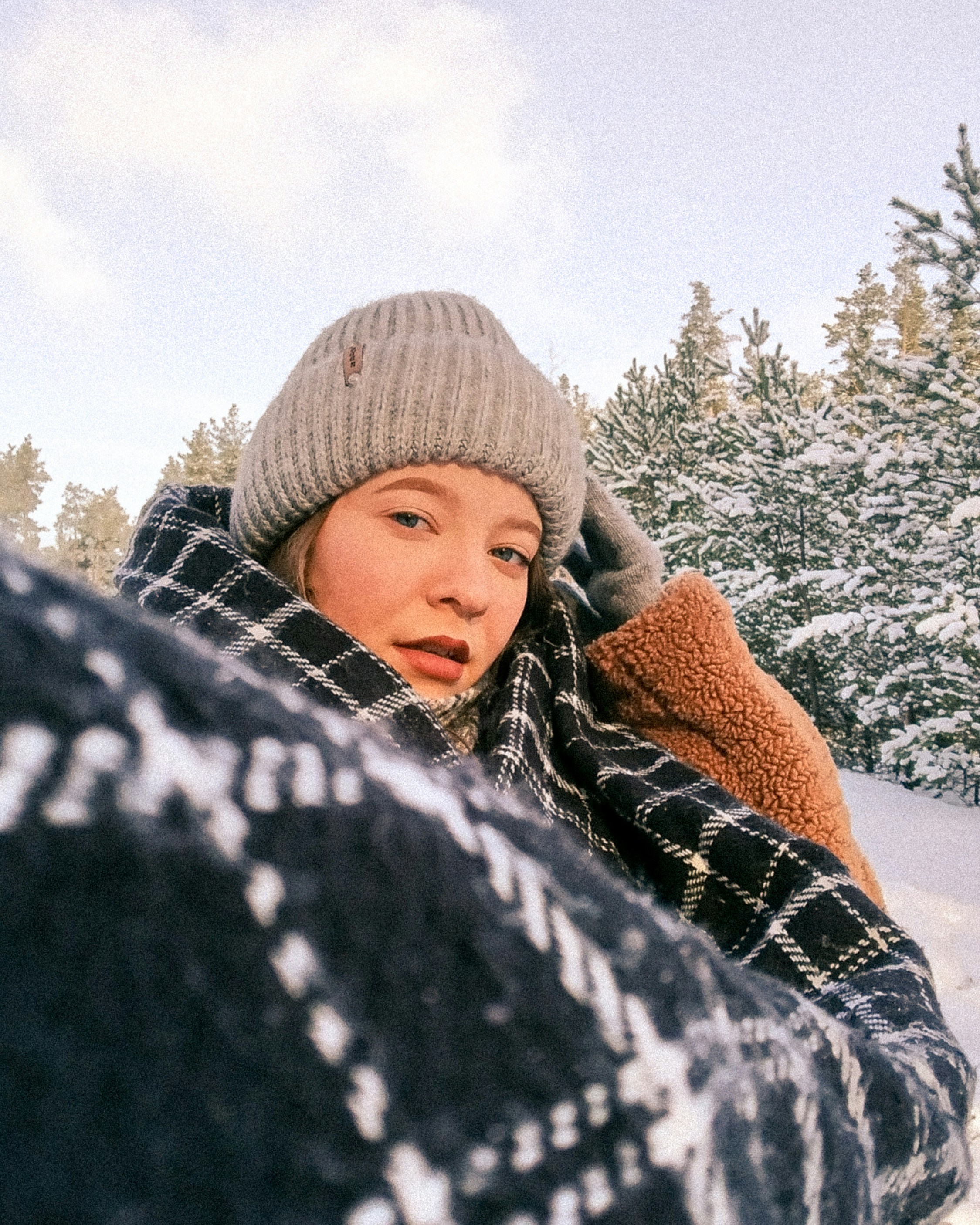Young person bundled in a plaid blanket, wearing a knitted hat, with a snowy forest backdrop.