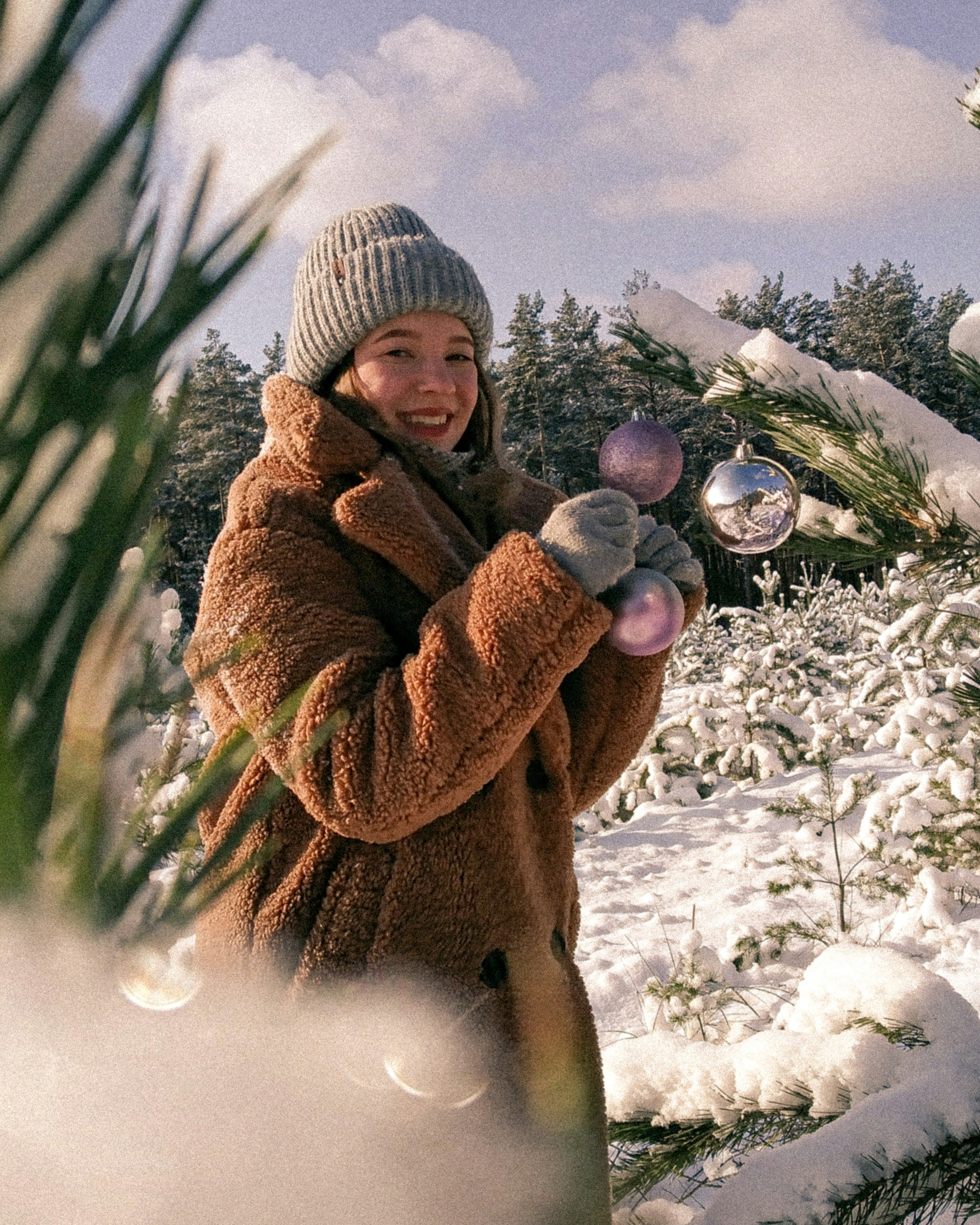 Photograph of a smiling woman in a brown teddy coat and gray beanie holding purple ornaments in a snowy forest under a clear sky.
