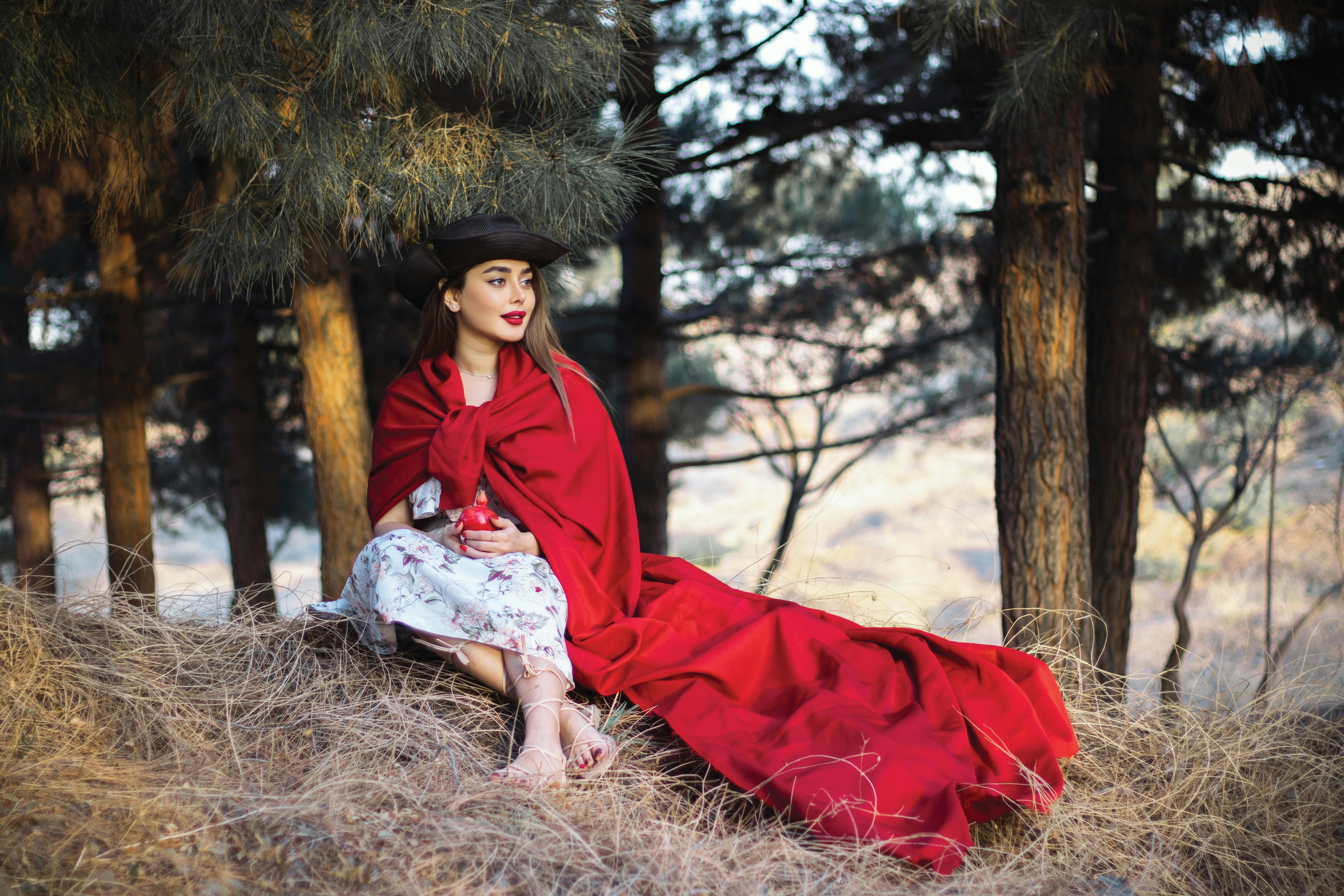 A young woman draped in a vibrant red blanket sits gracefully among tall pine trees, exuding a tranquil aura in a sunlit forest setting.