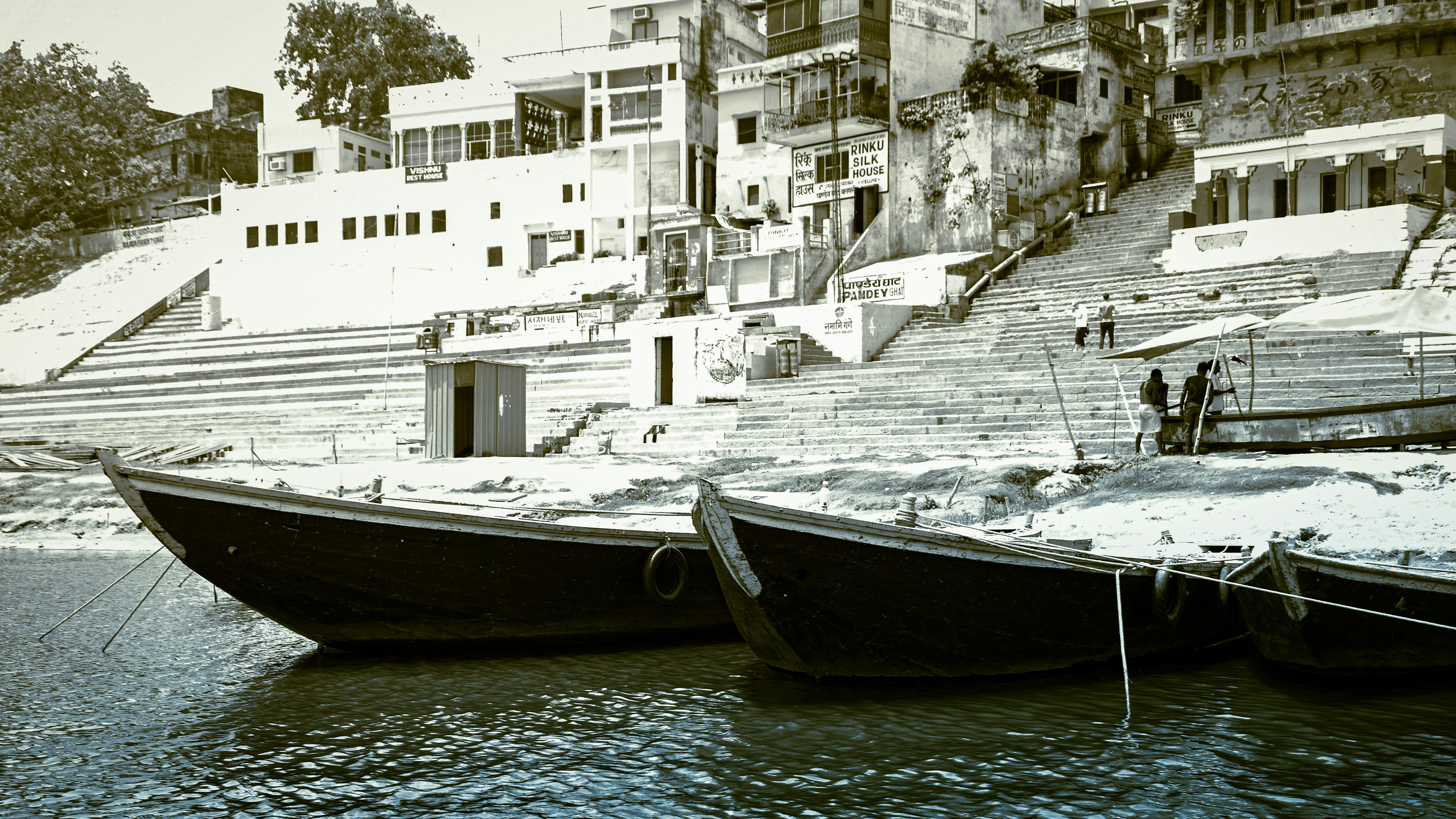 Two traditional boats anchored by a riverbank, with steps leading to a series of buildings in the background. The scene is captured in a monochrome aesthetic.
