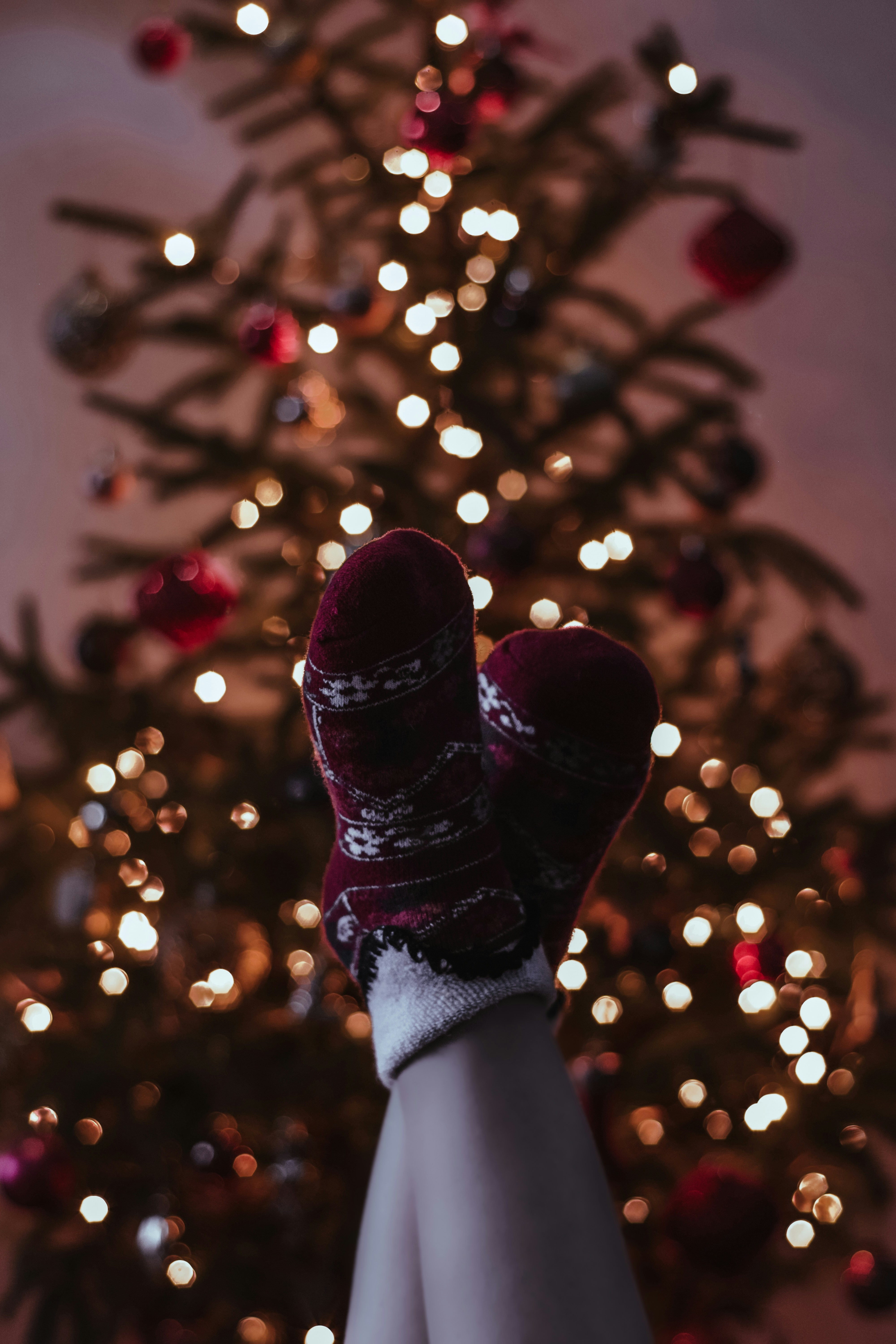 Feet in festive socks resting against a blurred Christmas tree adorned with twinkling lights and ornaments.