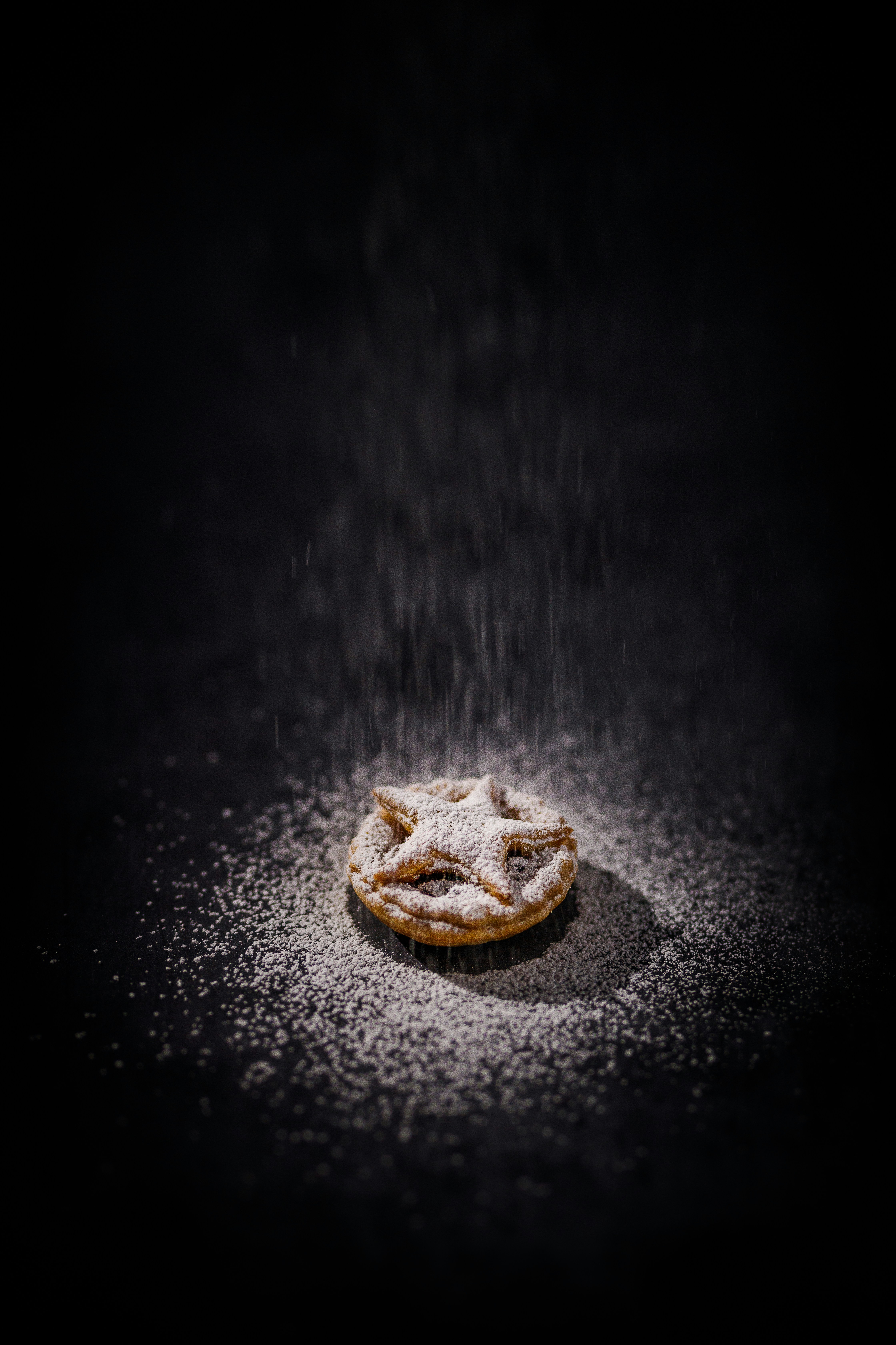 Star-shaped cookie resting on a dark surface, gently dusted with powdered sugar. The scene captures the delicate process of adding sweetness.