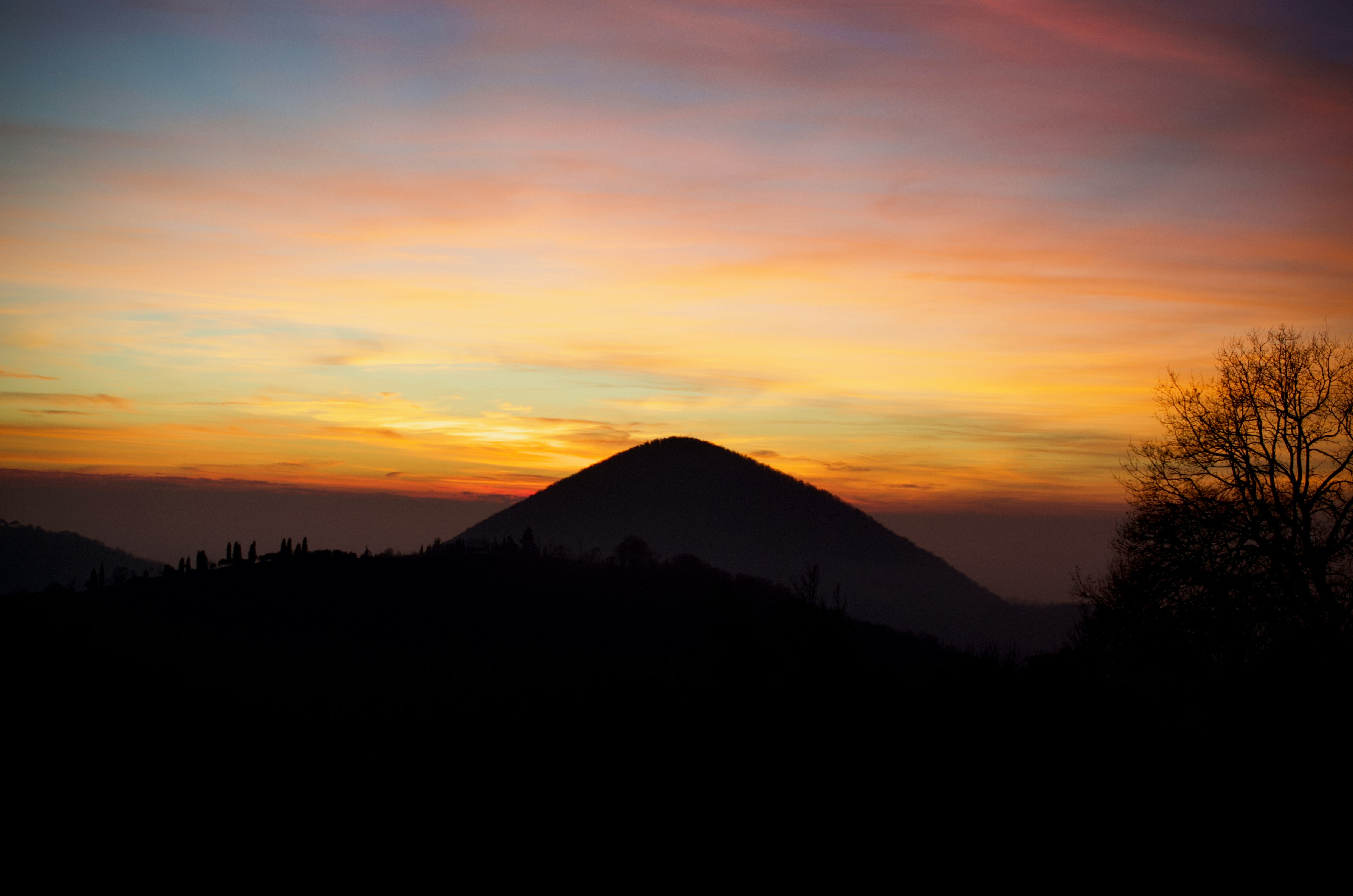 Silhouette of a mountain against a vibrant twilight sky, with soft hues transitioning from orange to blue. A solitary tree stands to the right, adding depth to the serene landscape.