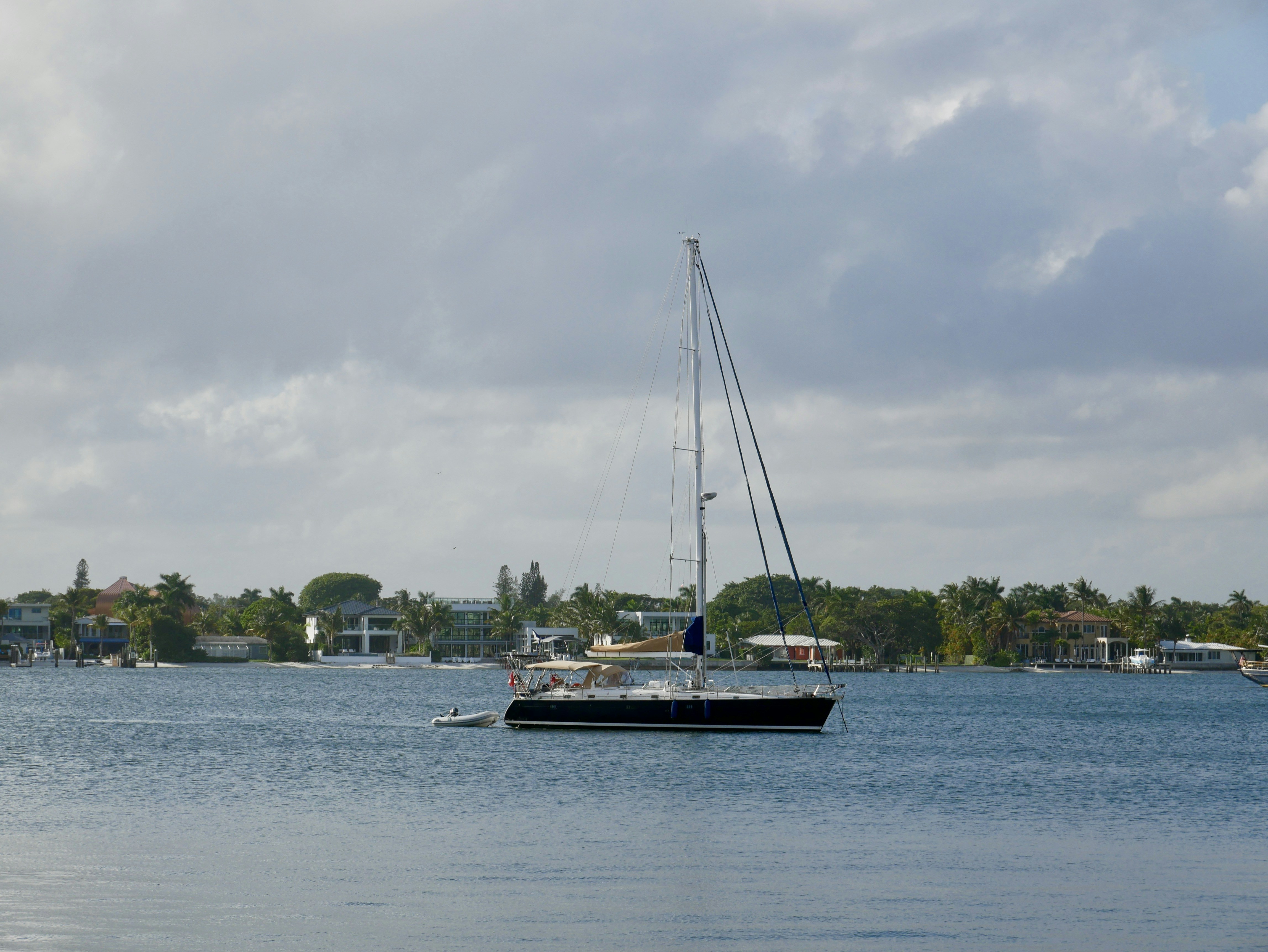 Sailboat gliding across calm waters, surrounded by lush waterfront properties under a cloudy sky.