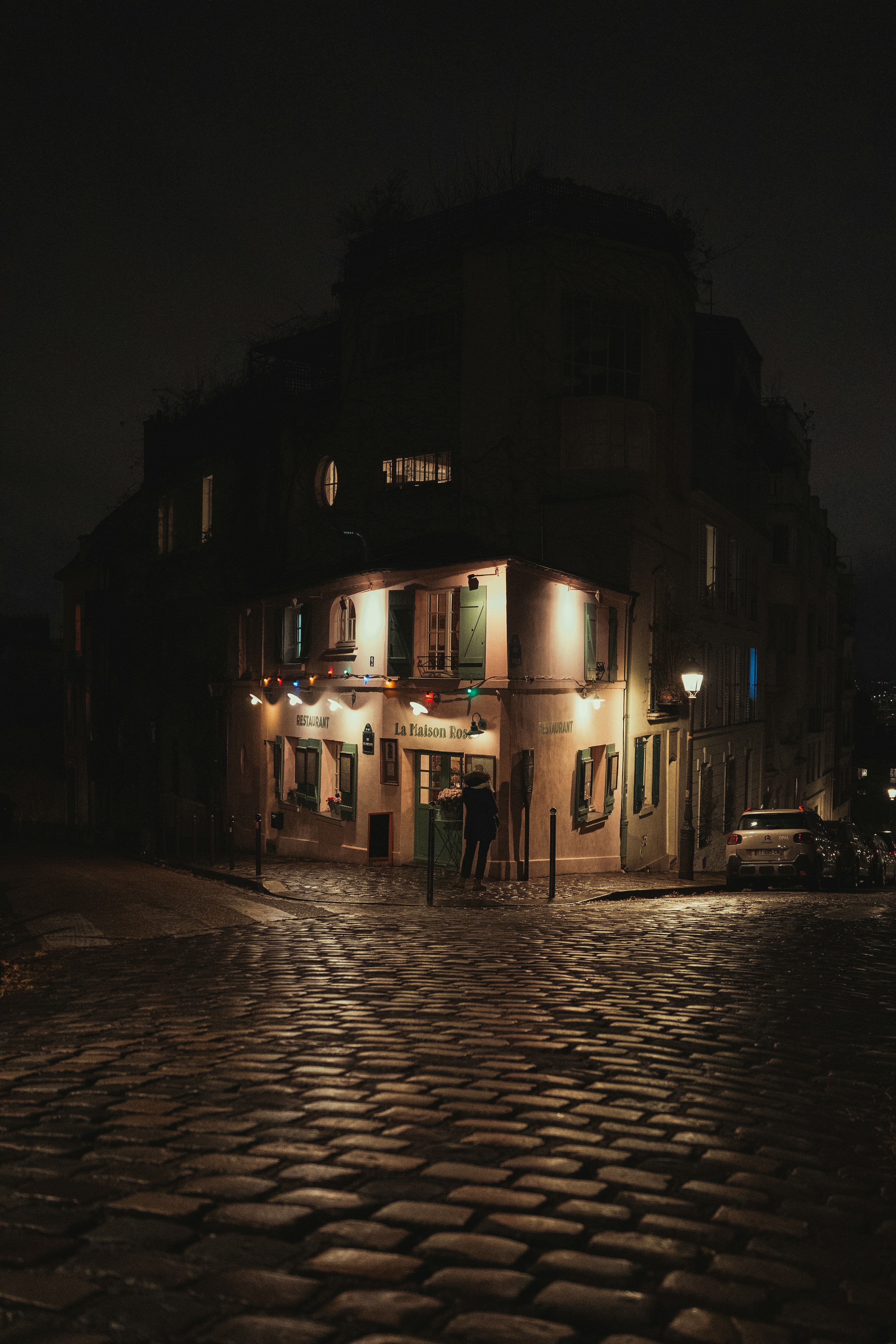 Charming street scene featuring a quaint restaurant illuminated by warm lights, nestled among cobblestone paths under a night sky.