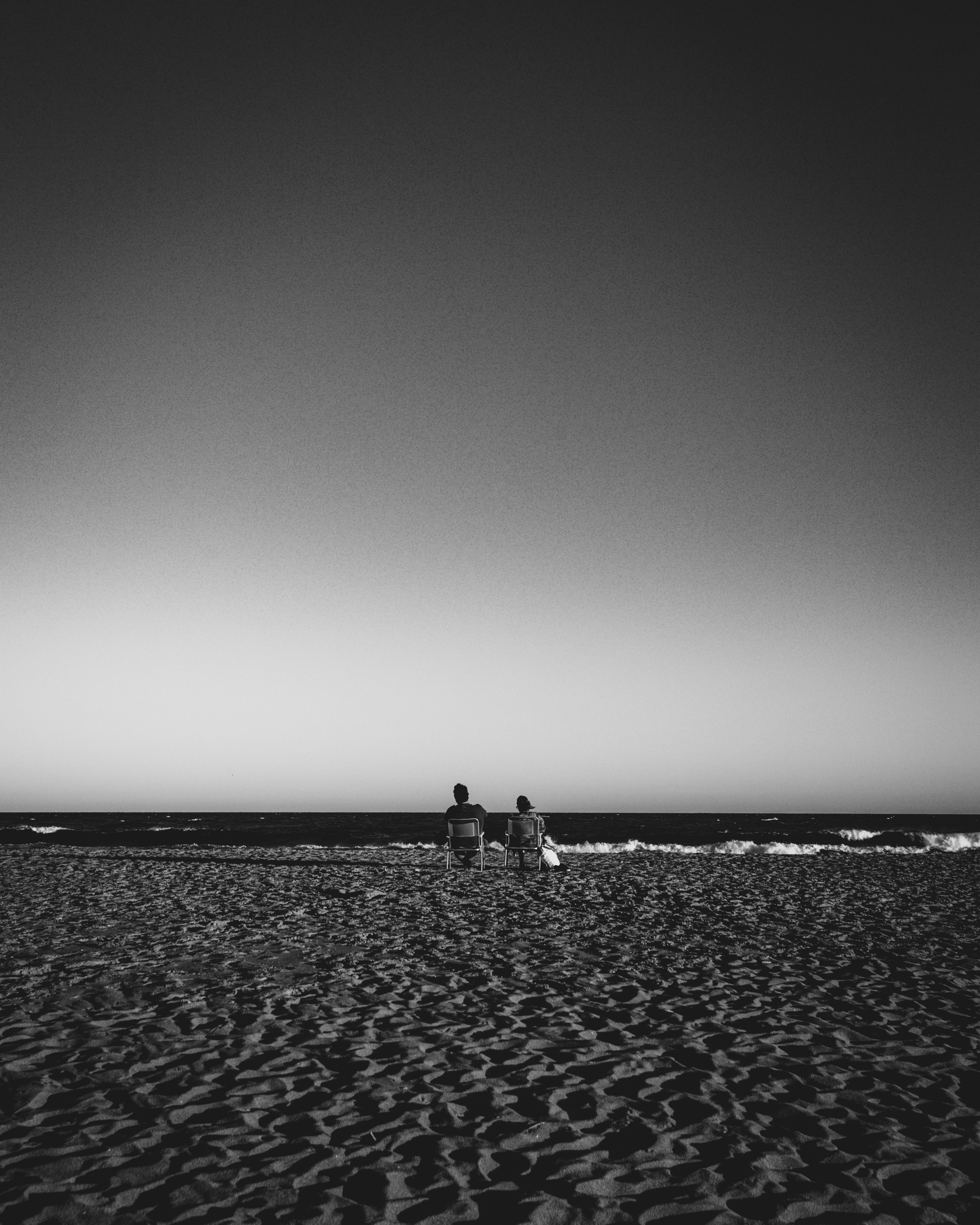Two figures seated on a beach, gazing at the horizon where the ocean meets the sky in monochrome tones.
