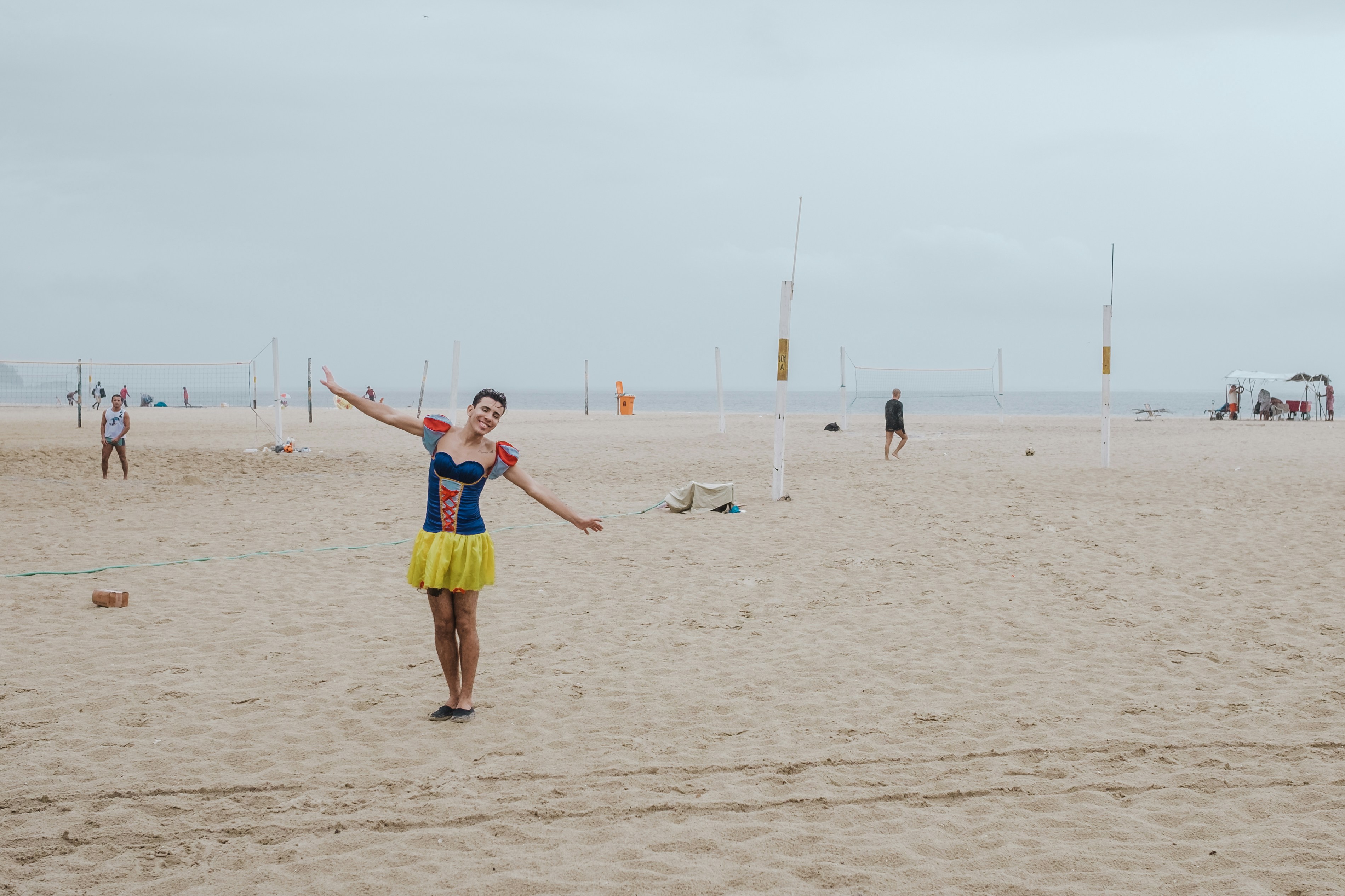 A joyful figure in a colorful costume dances on a sandy beach, surrounded by a serene coastal landscape. The scene captures a playful moment amidst a calm atmosphere.
