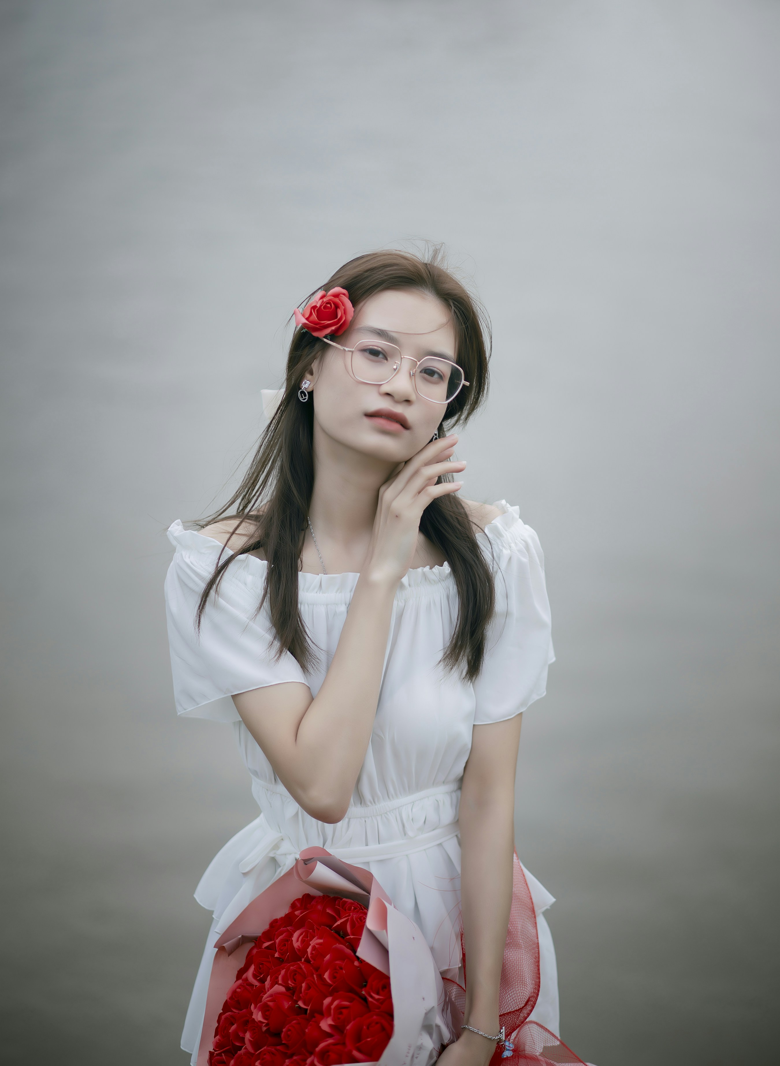 Young woman in a white dress with a rose in her hair, holding a bouquet of red flowers against a muted background.