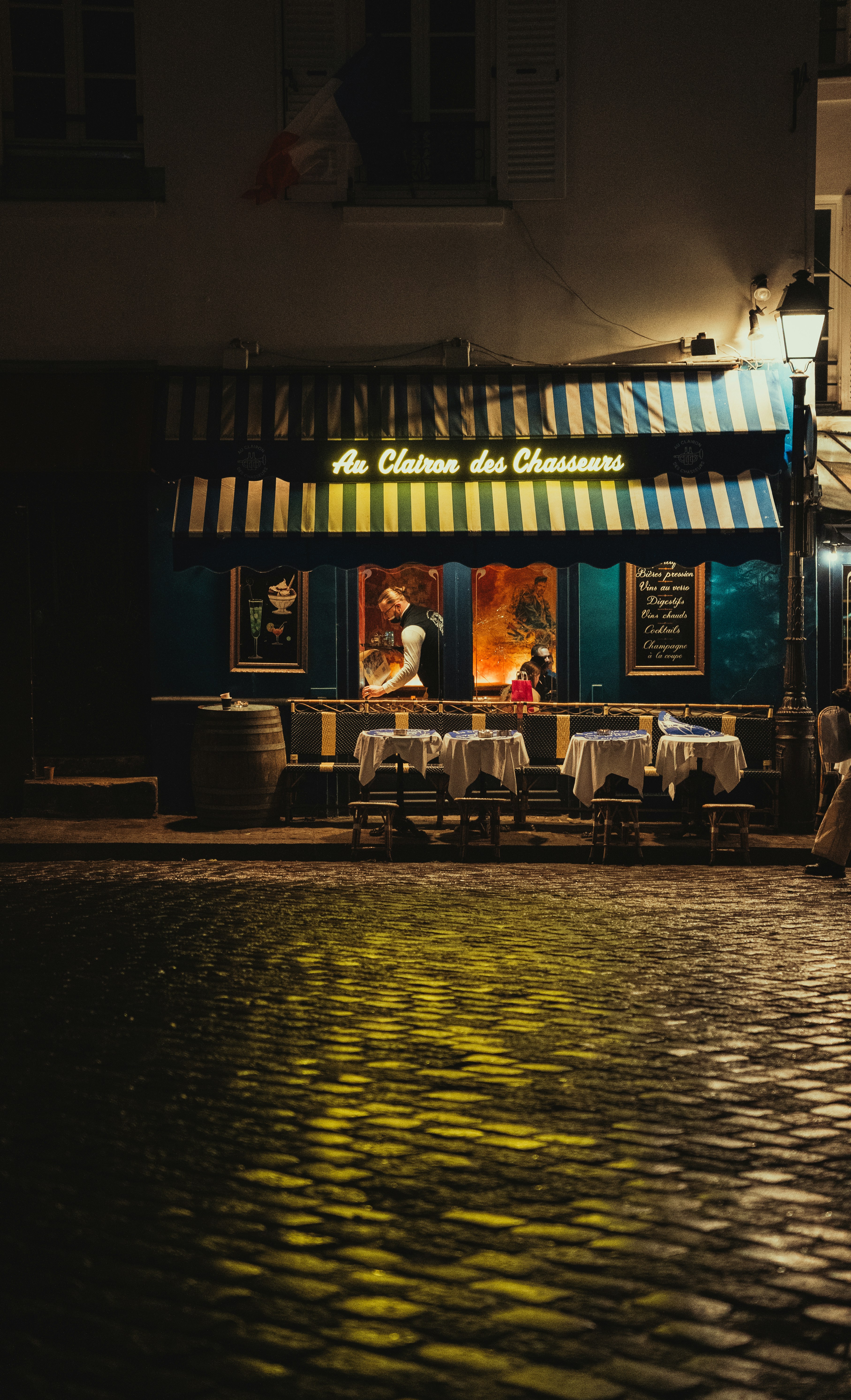 Dimly lit Parisian café with tables set under a striped awning, reflecting warm light on cobblestone street.