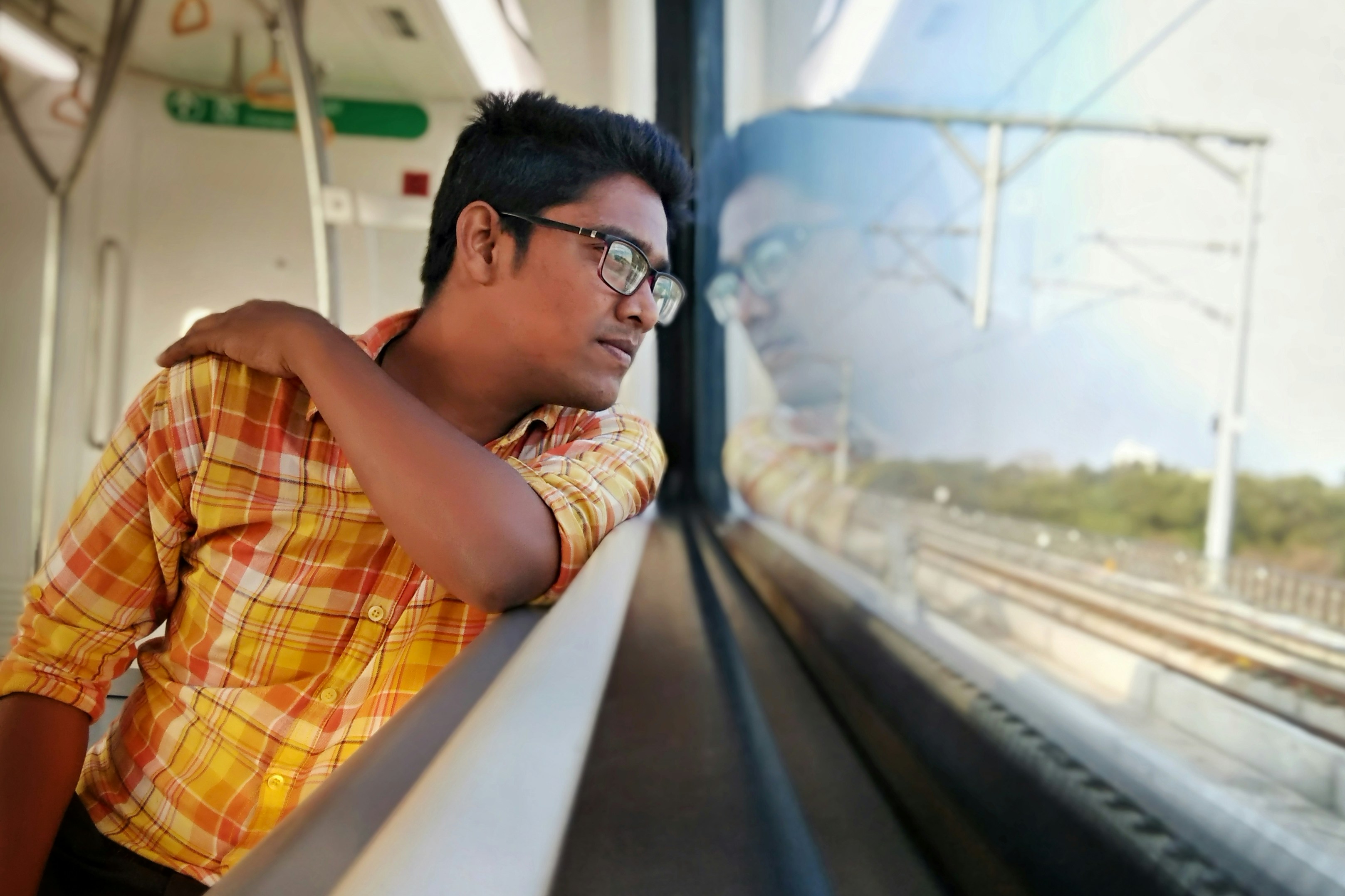 A young man gazes out the window of a train, lost in thought, with reflections of the landscape visible in the glass.