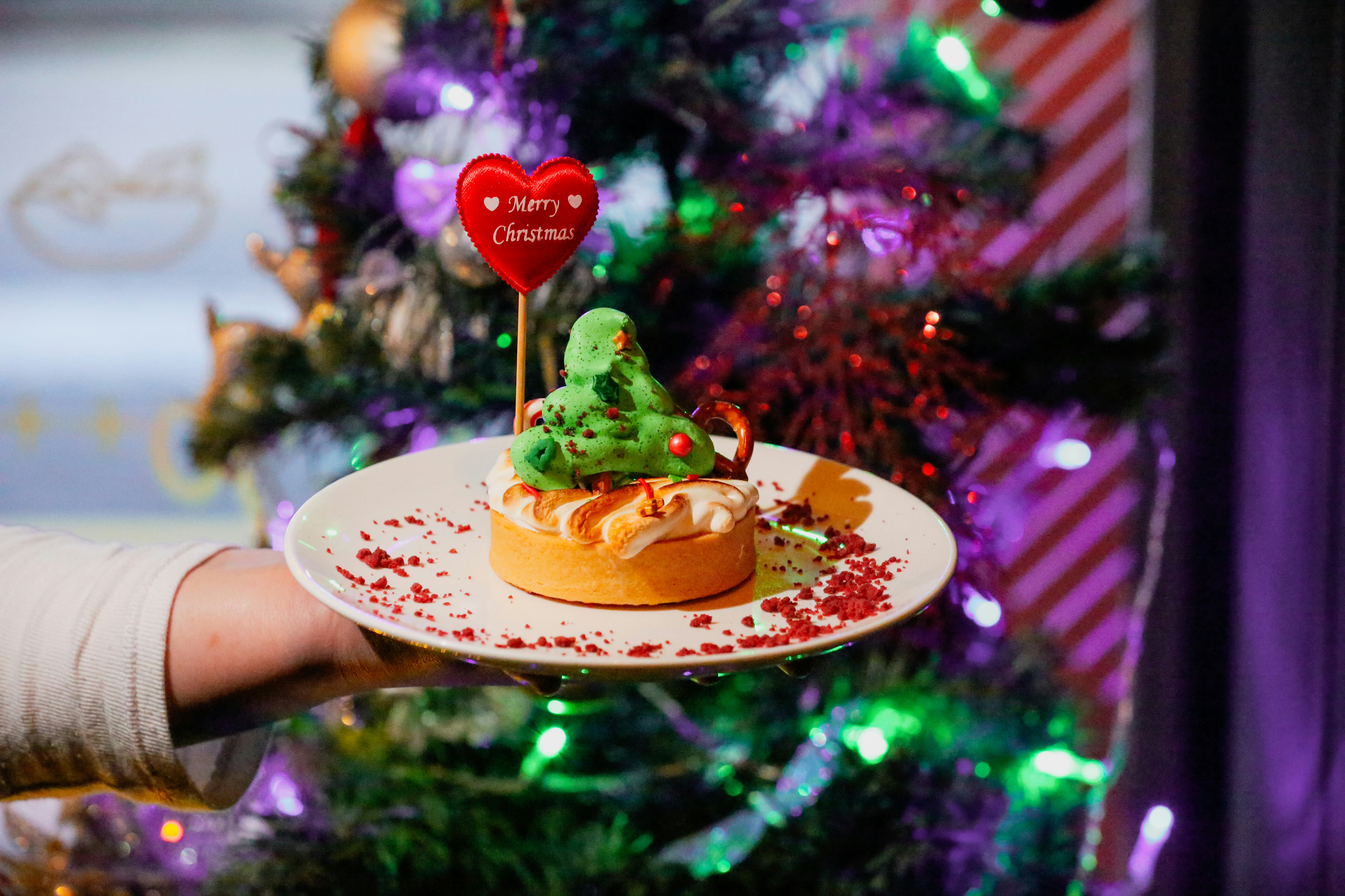 A holiday-themed dessert featuring a green creature atop a creamy tart, presented against a backdrop of a decorated Christmas tree.