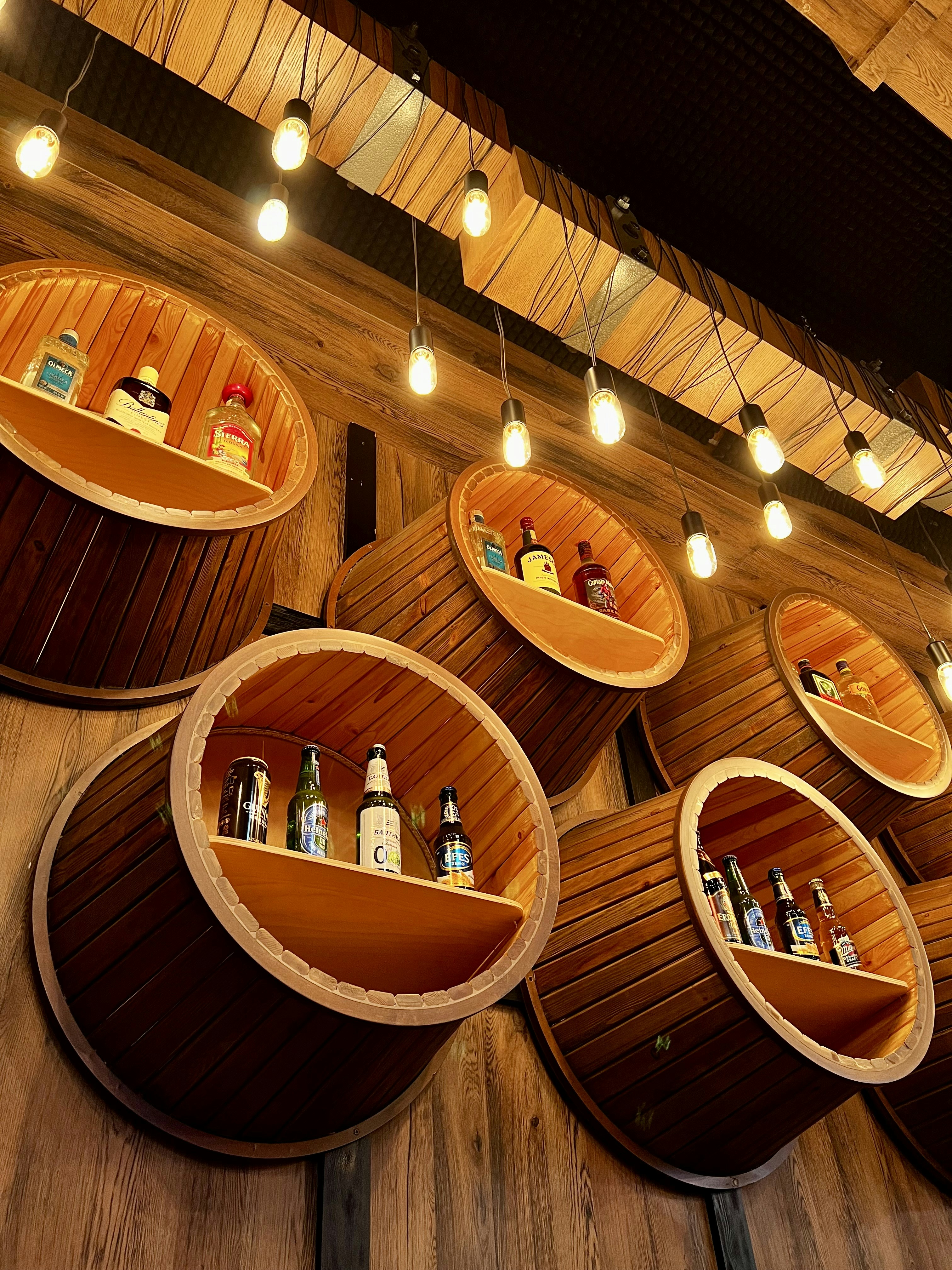 Circles of Light: Bottled Shelves on a Barrel-Wall Under Edison Glow