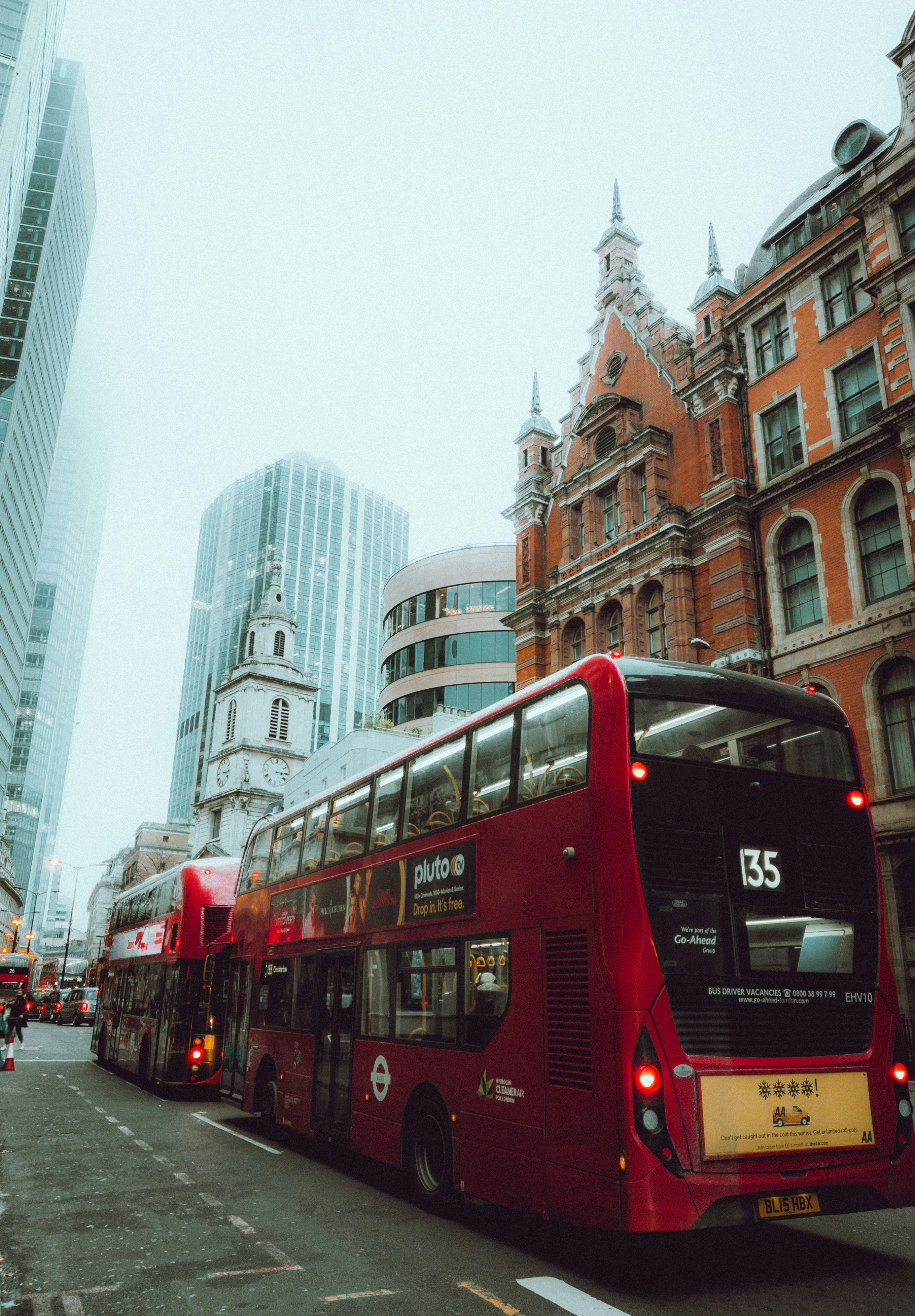 Red double-decker bus on a foggy urban street flanked by ornate brick buildings and modern glass towers.