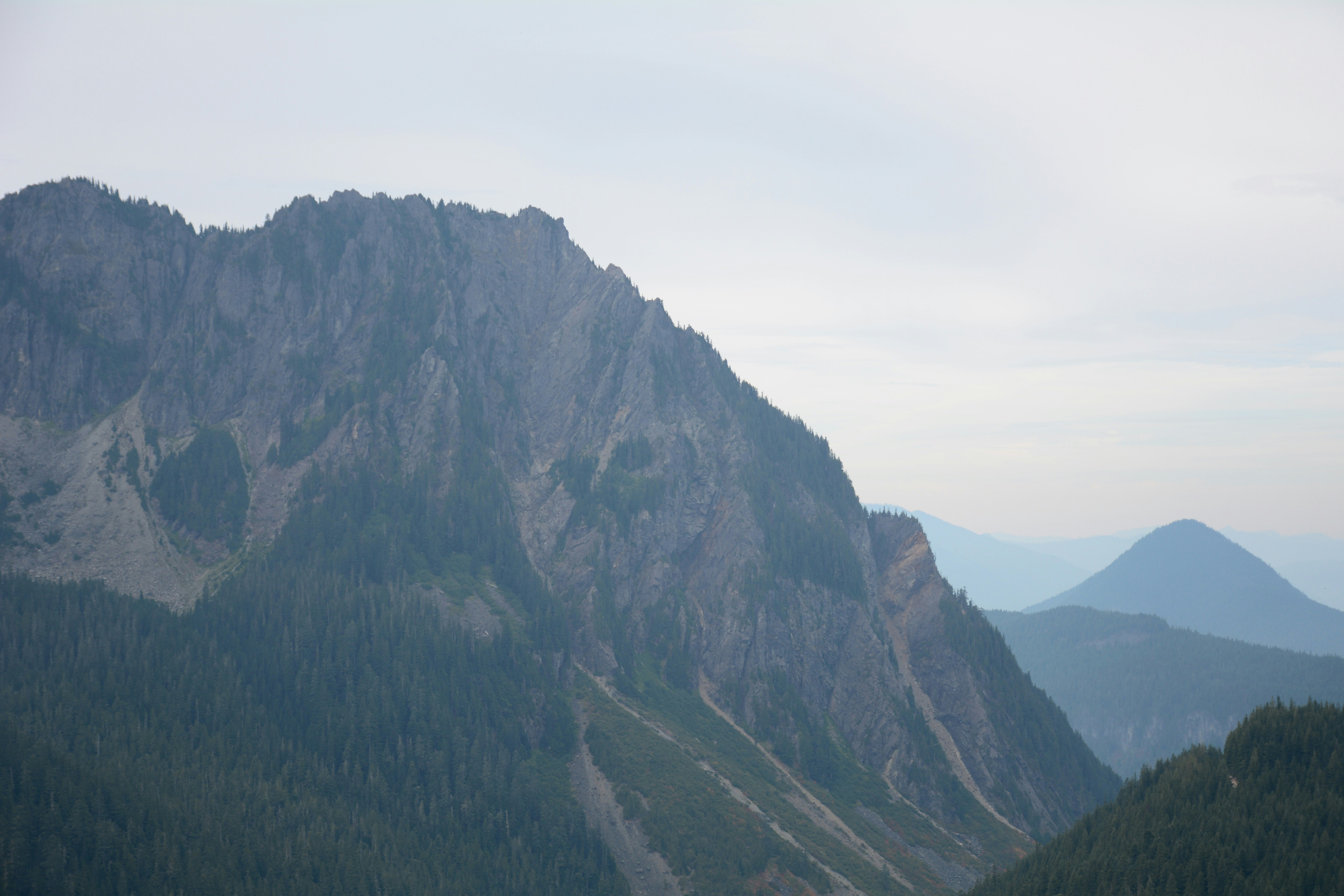 Majestic mountain range with rugged peaks and lush green forests under a soft, overcast sky.