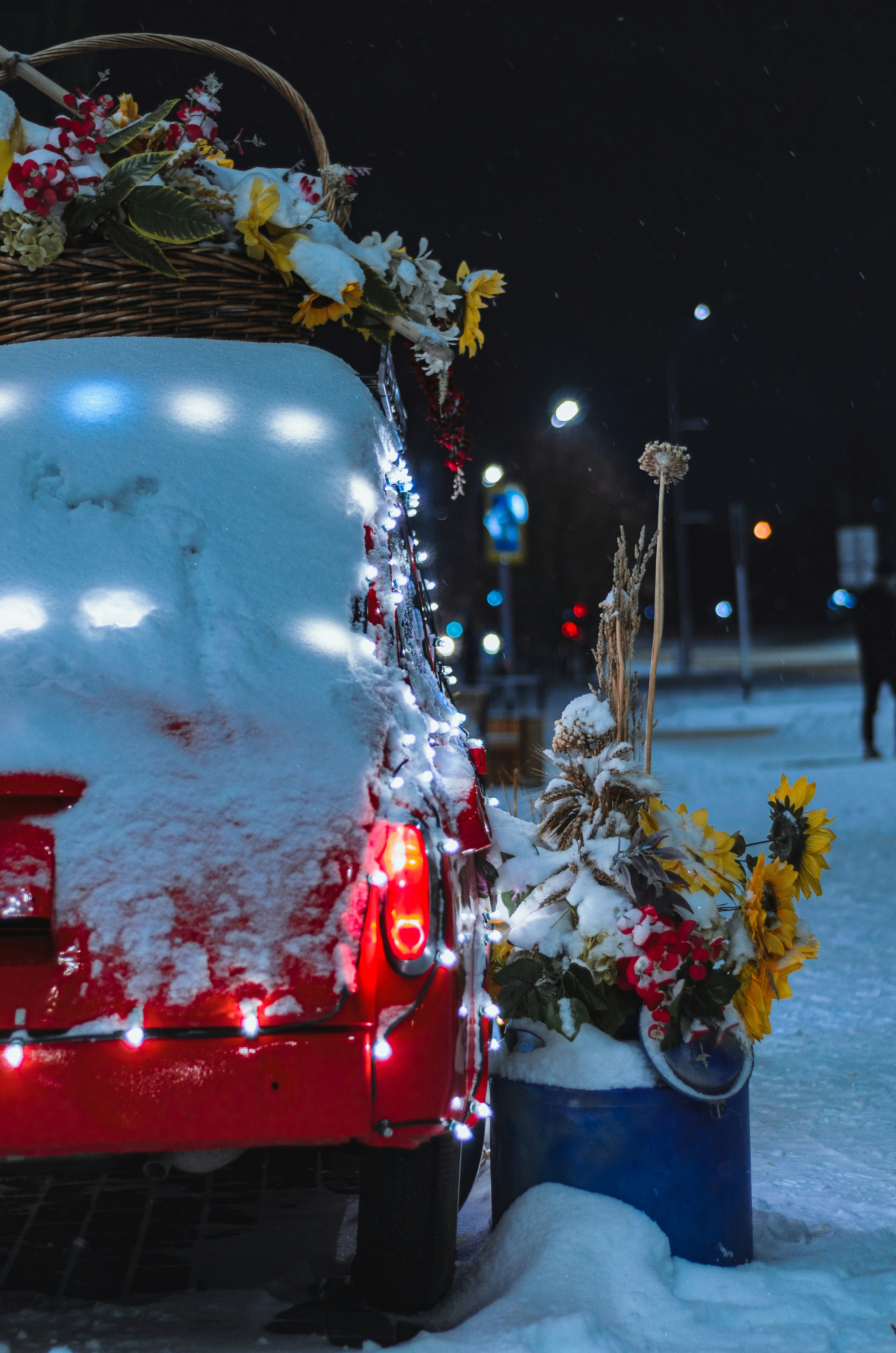 A vintage red car adorned with snow and twinkling lights, surrounded by colorful flowers in a bucket. The scene captures the charm of winter festivities.