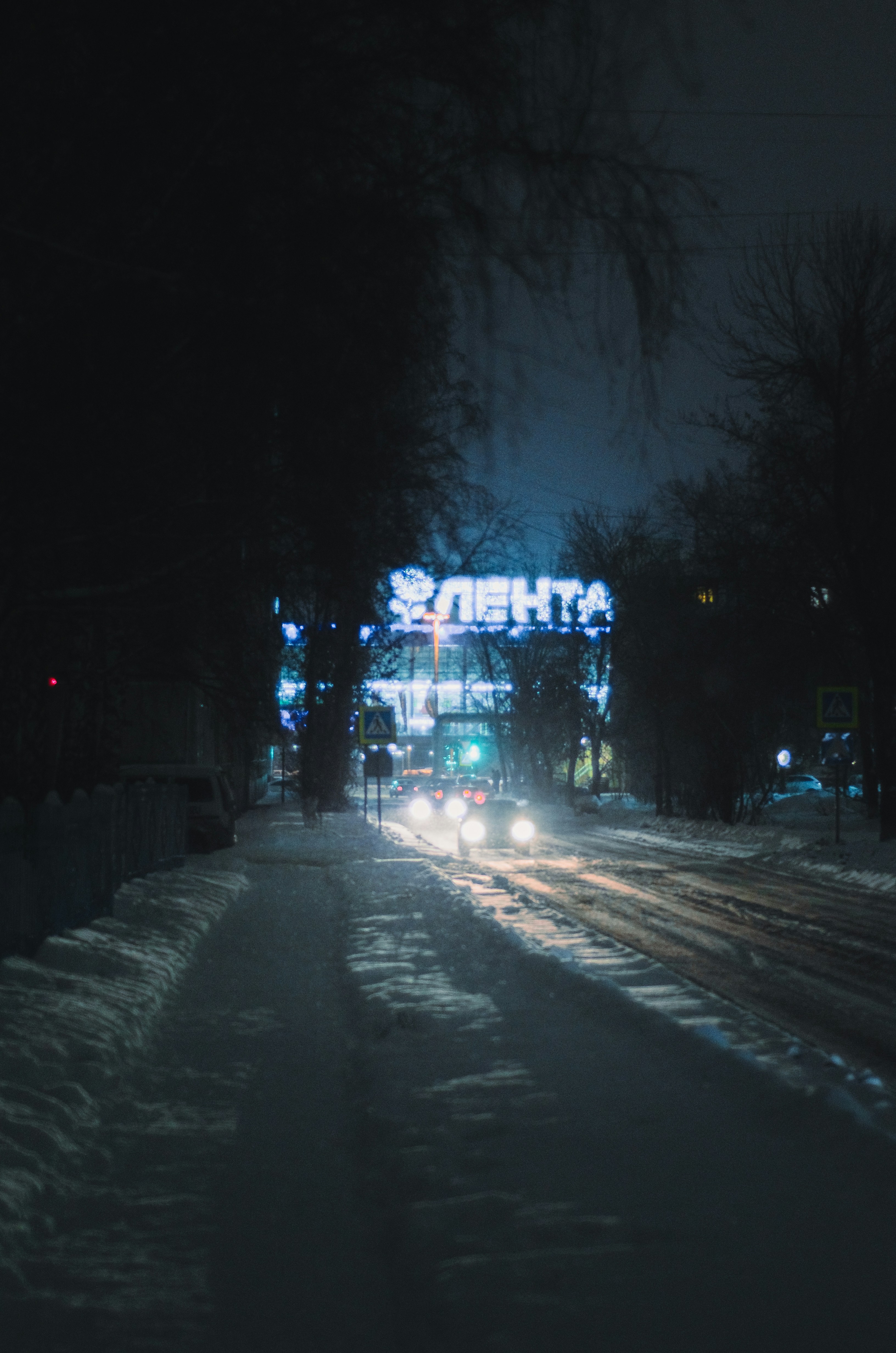 Illuminated storefront sign shines through a snowy street at night, with cars navigating the wintry conditions. 