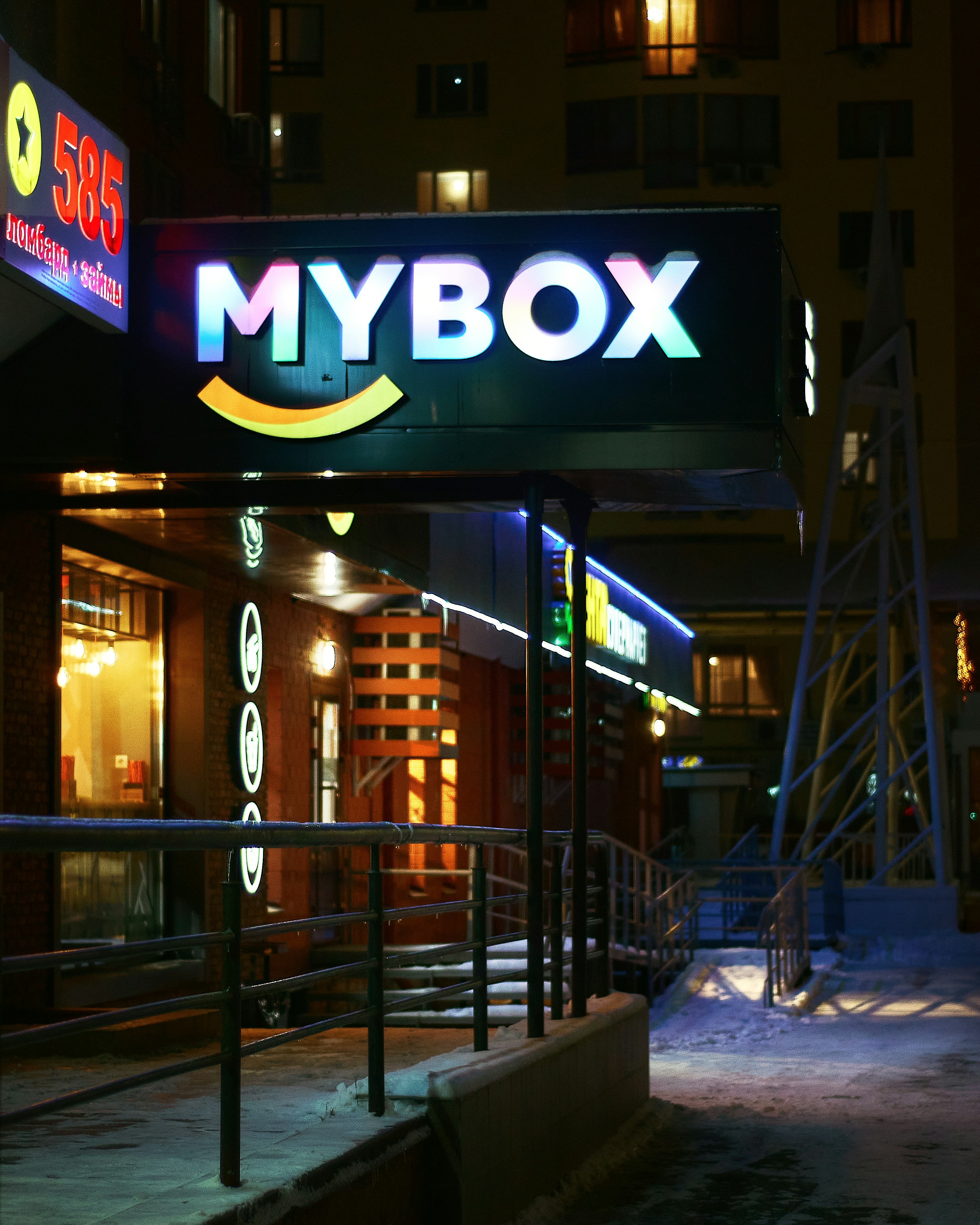 Vibrant MYBOX sign illuminated against a nighttime backdrop, showcasing modern architecture and inviting ambiance. The scene captures the essence of urban life after dark.