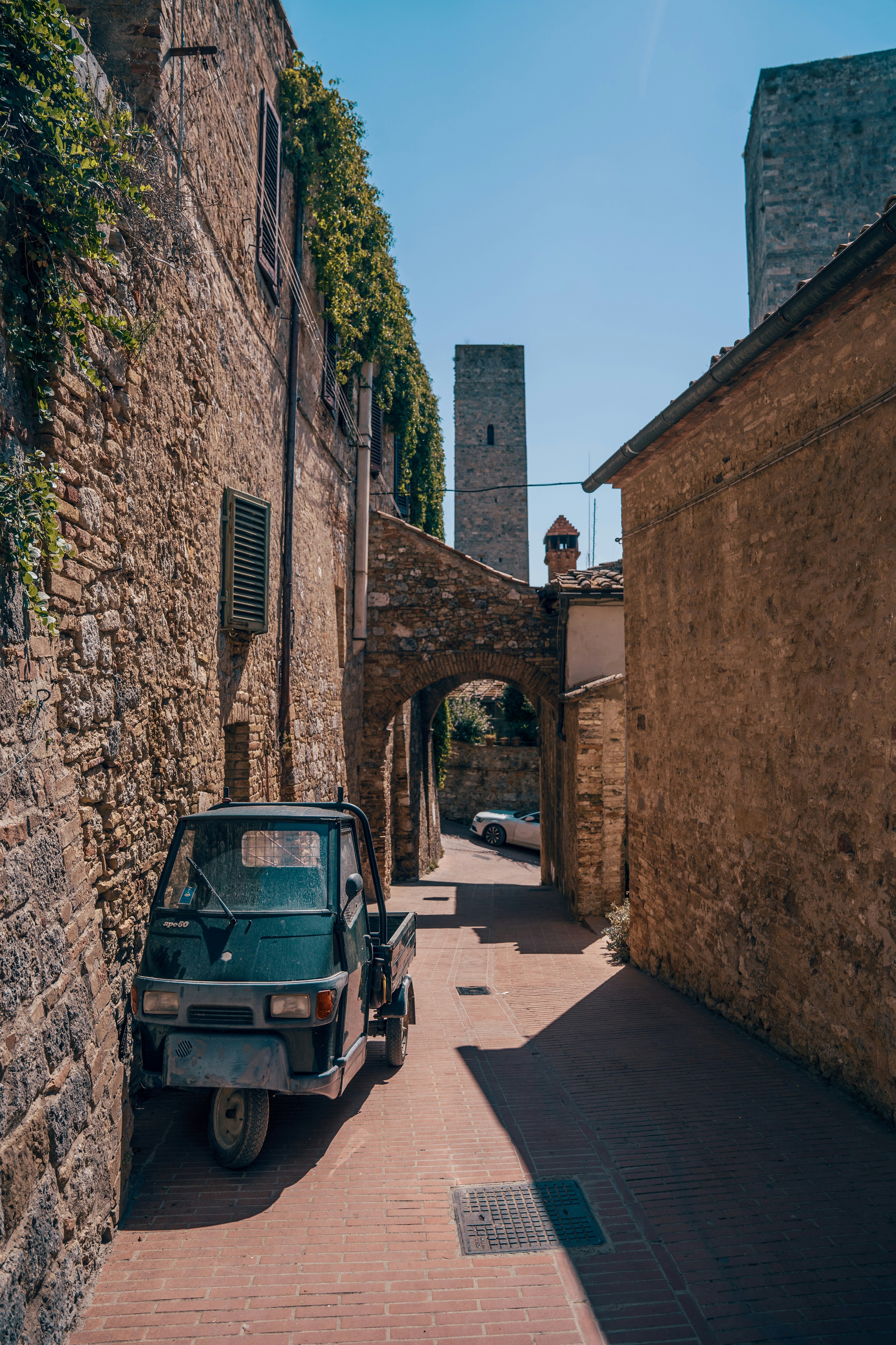 Narrow cobblestone alley lined with rustic stone walls, featuring a vintage green vehicle parked beside an archway. Sunlight casts soft shadows along the path.