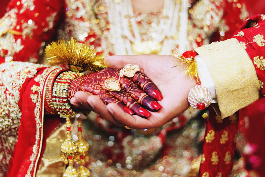 Close-up of a bride’s mehndi-decorated hand resting in the groom’s palm, with red and gold wedding attire filling the background.