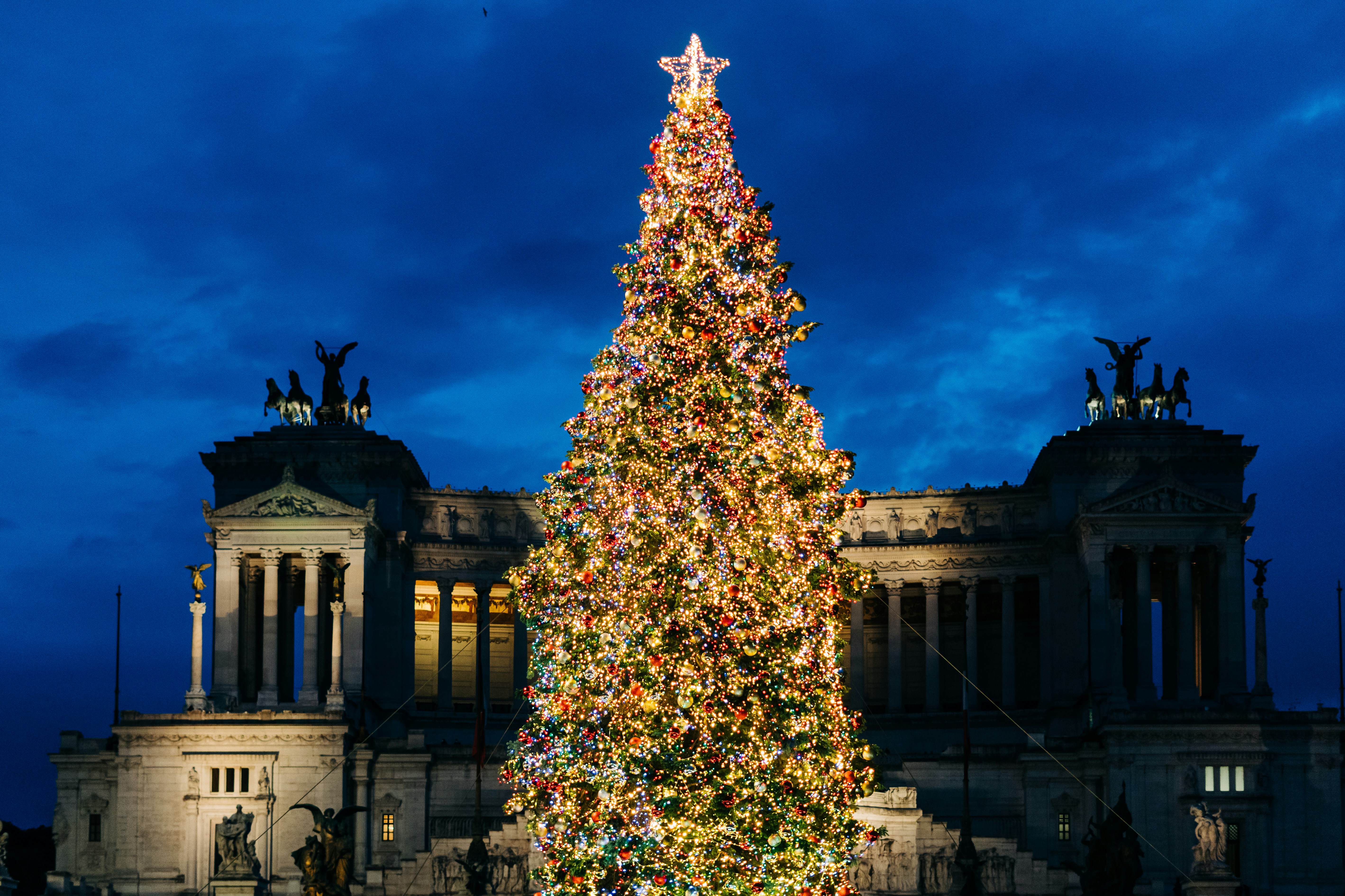 A vibrant Christmas tree adorned with colorful lights stands prominently in front of a grand historical building, illuminated against a twilight sky.