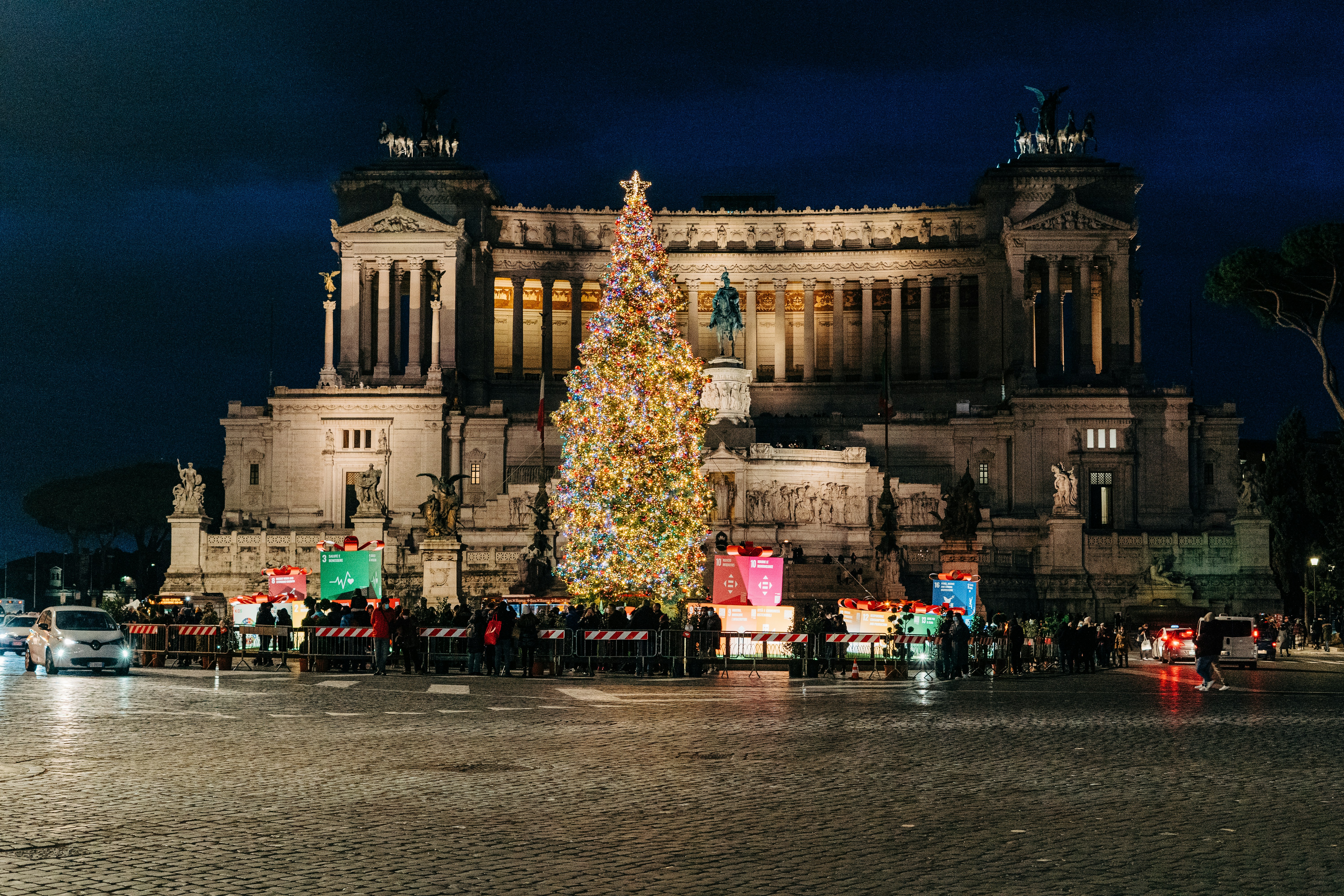 A vibrant Christmas tree adorned with colorful lights stands in front of the historic Altare della Patria, bustling with holiday cheer and visitors. 