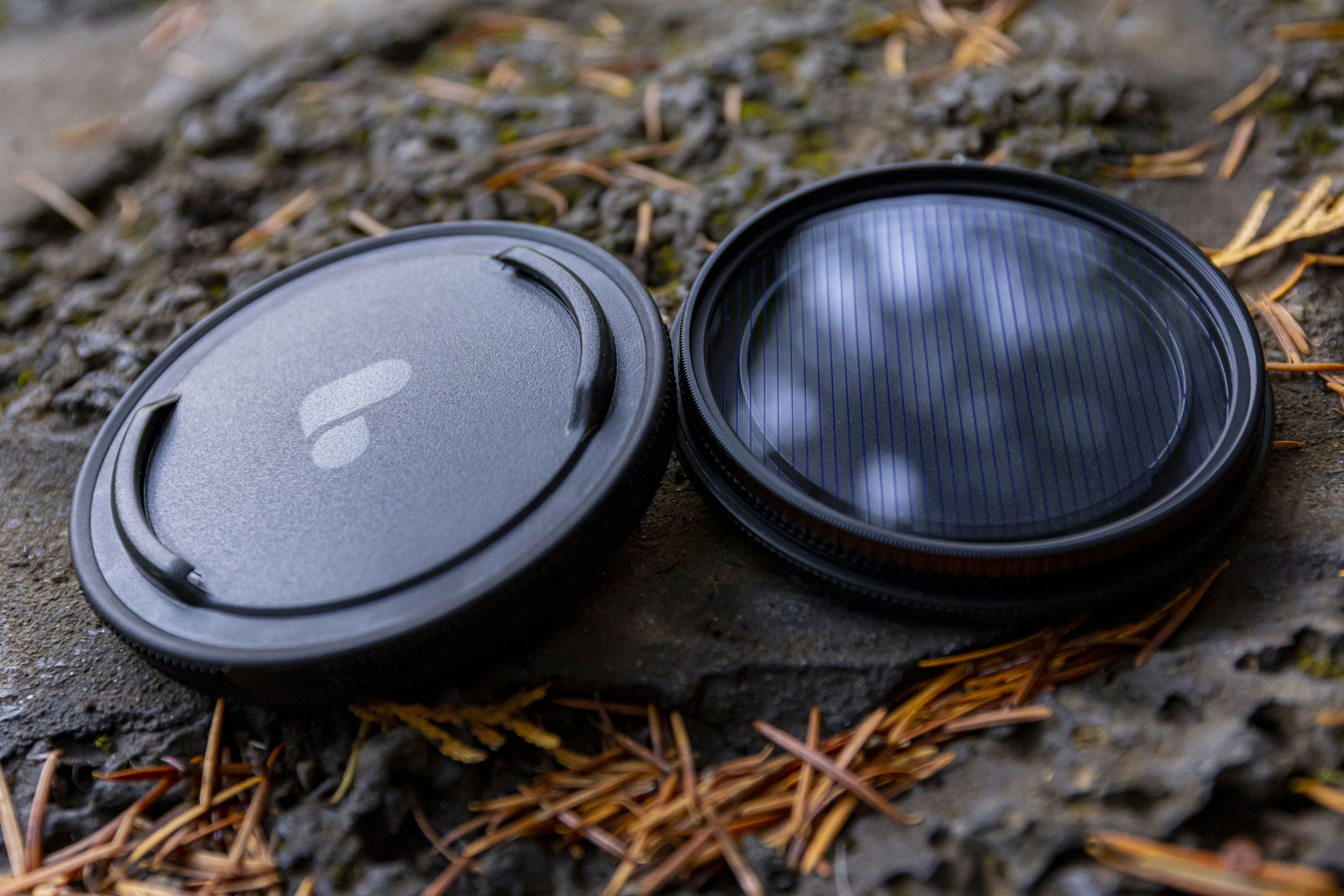 Two lens caps lie on a textured rock surface surrounded by scattered pine needles, showcasing a blend of nature and photography gear.