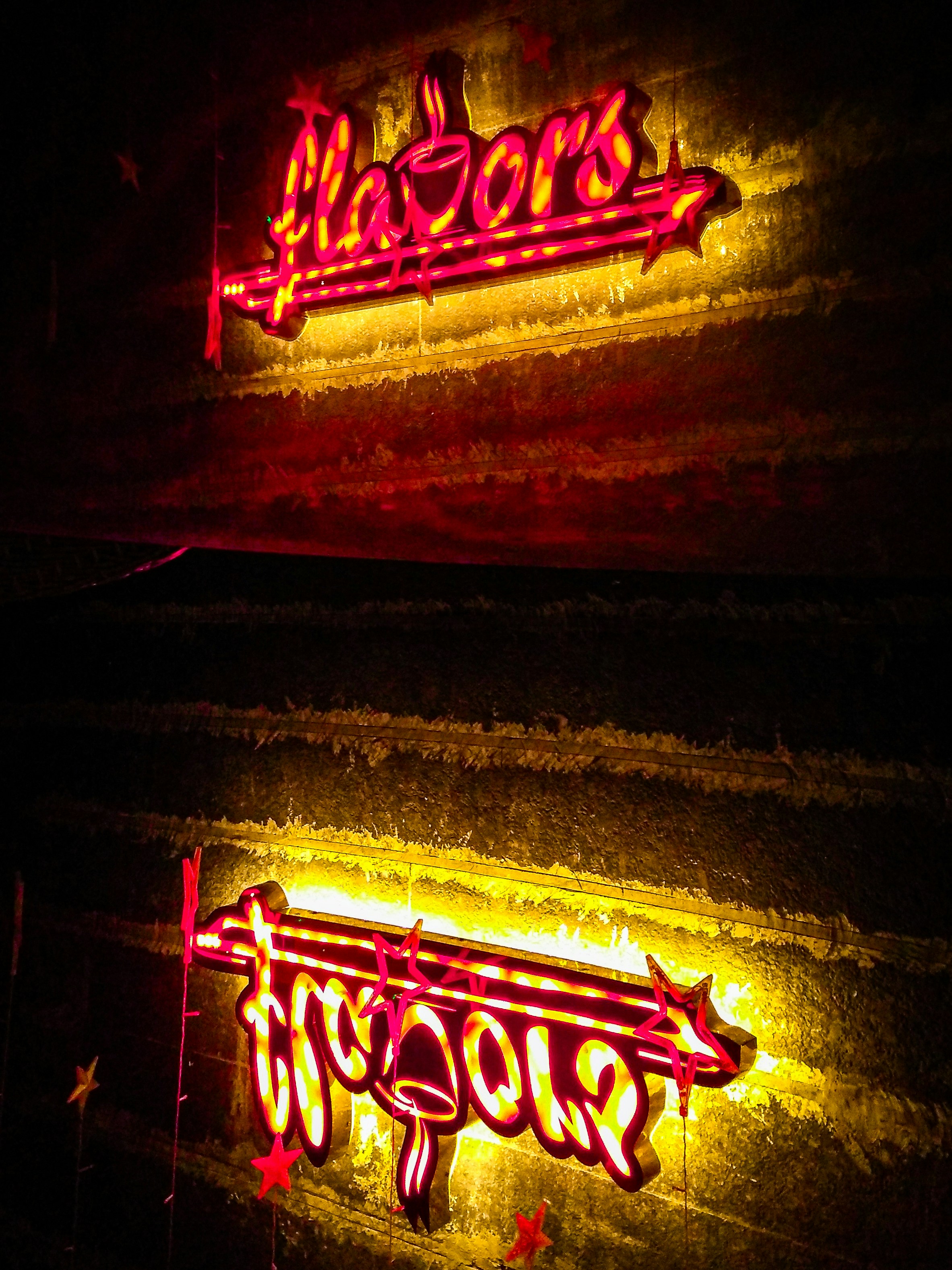 Neon pink sign atop a weathered wall, with a warm yellow reflection shimmering on a rain-wet street.