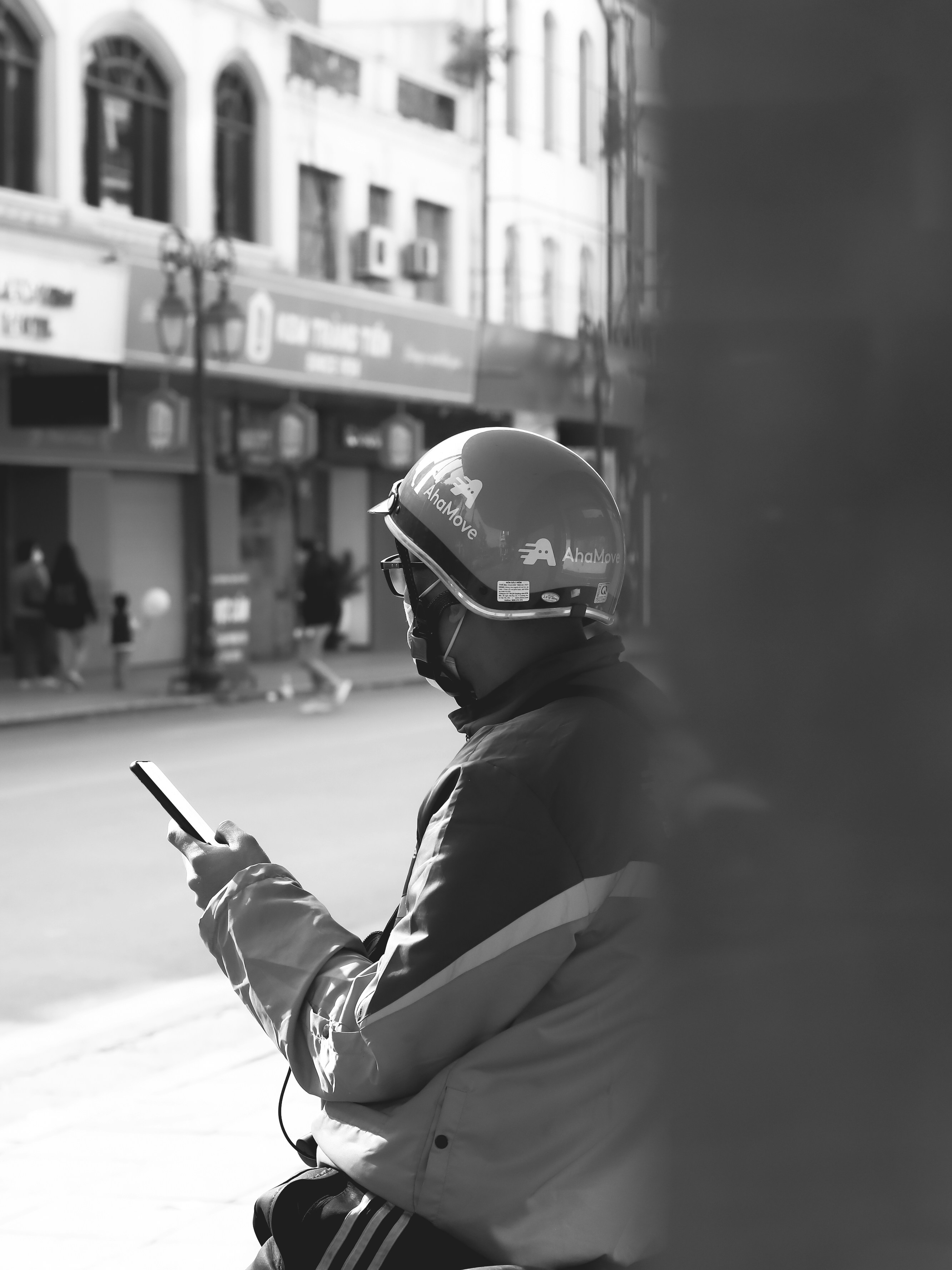 Delivery rider checking a mobile device while seated against a wall in a bustling urban environment.