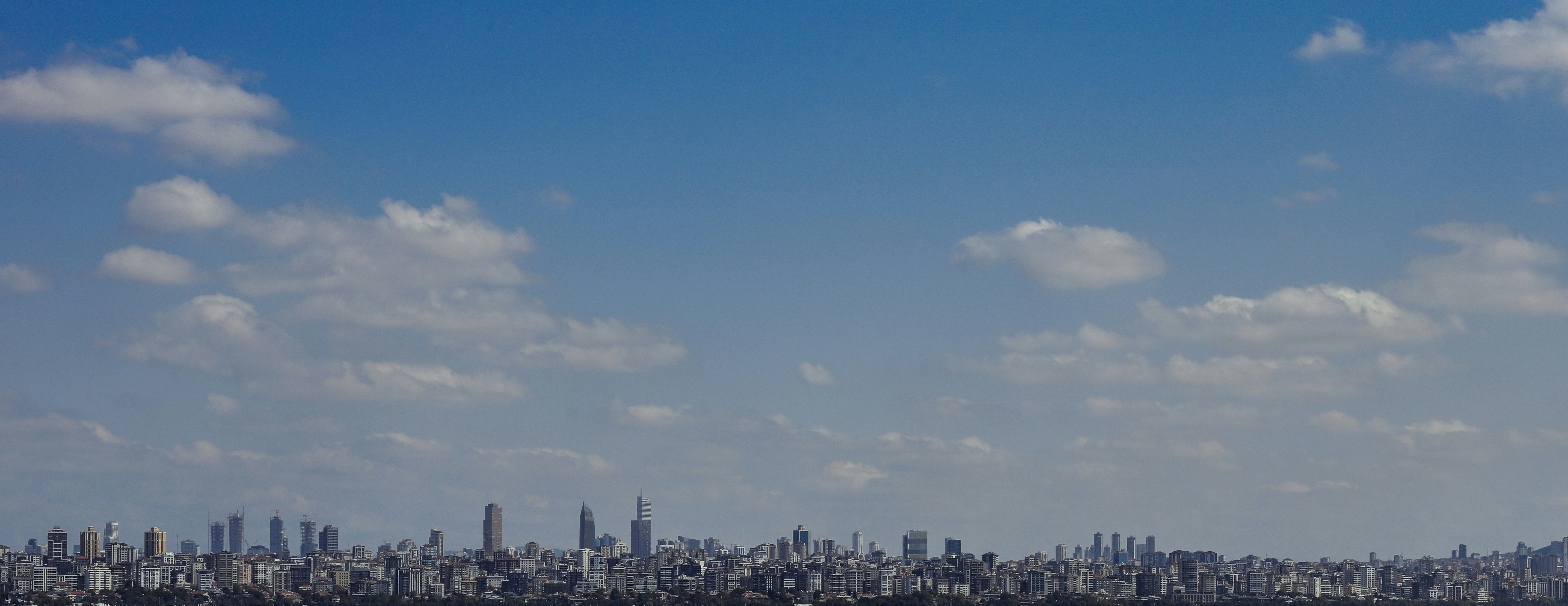 A panoramic view of a sprawling city skyline against a backdrop of fluffy clouds and a bright blue sky.