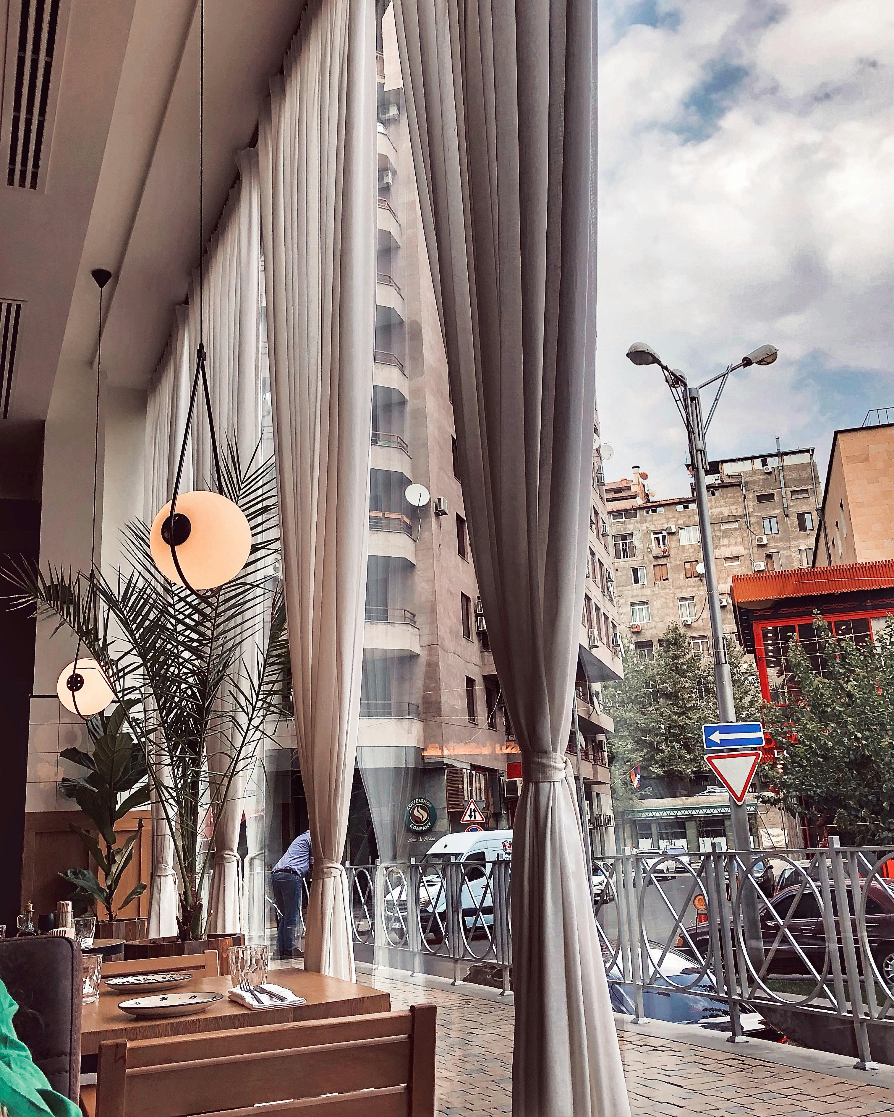 Chic restaurant interior featuring elegant curtains and modern lighting, with a view of bustling city life outside. 