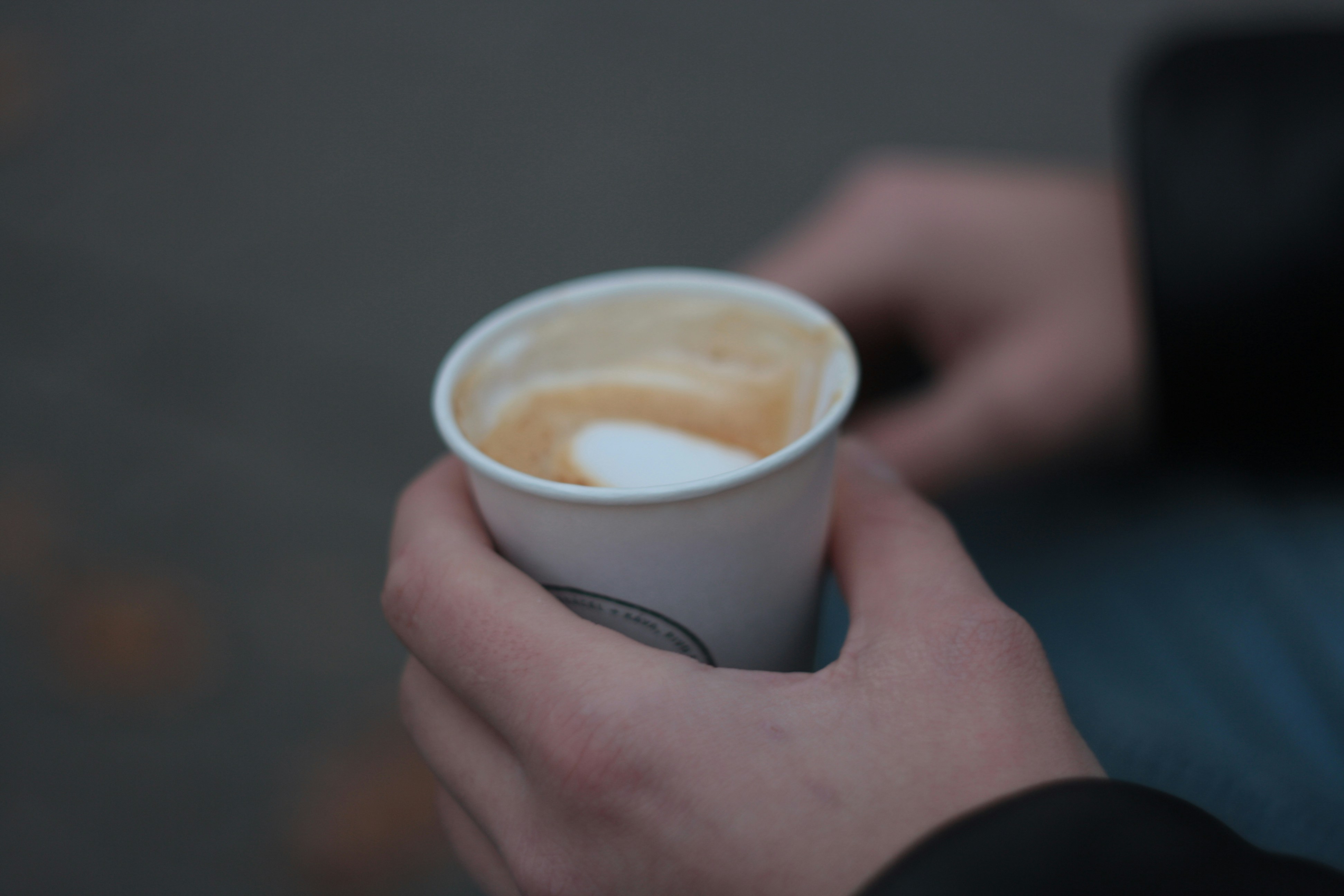 A hand holding a cup of coffee, showcasing the creamy texture and frothy top against a blurred background. The scene evokes a sense of warmth and relaxation.