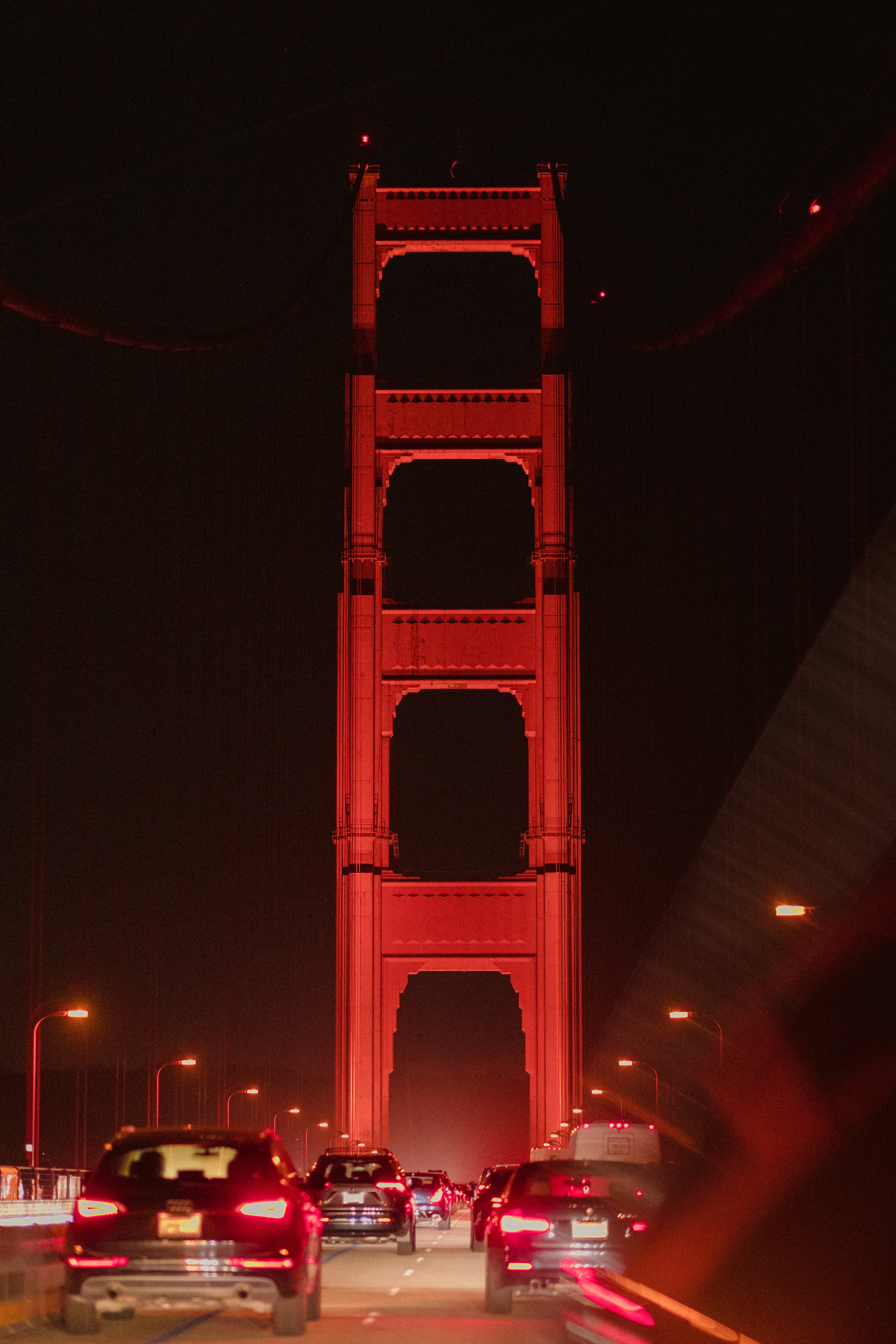 Golden Gate Bridge towering over traffic at night, illuminated in vibrant red hues. Cars line the roadway beneath the bridge's majestic structure.
