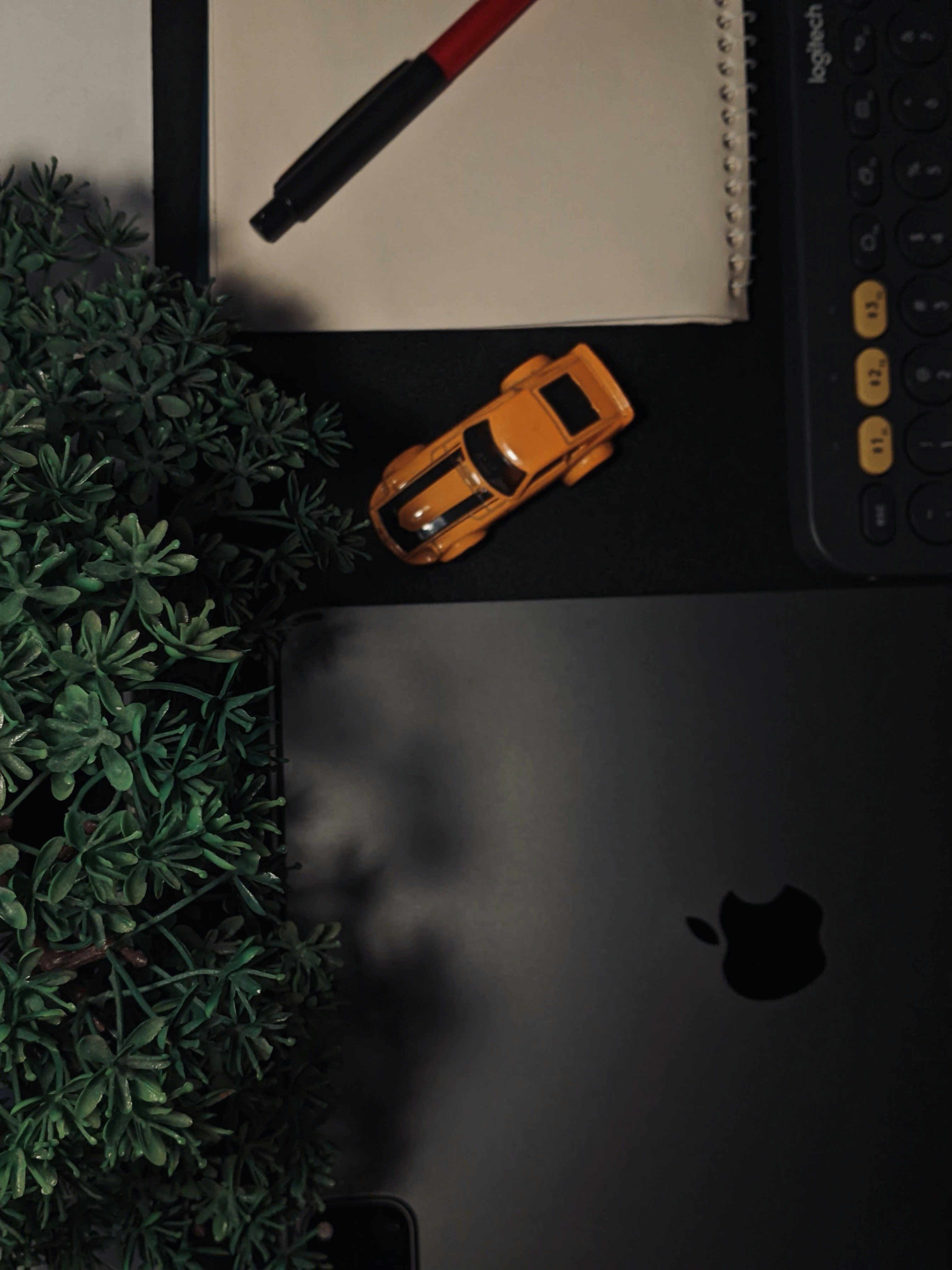 Orange Toy Car on Dark Desk Amid Green Foliage and Apple Logo Tablet
