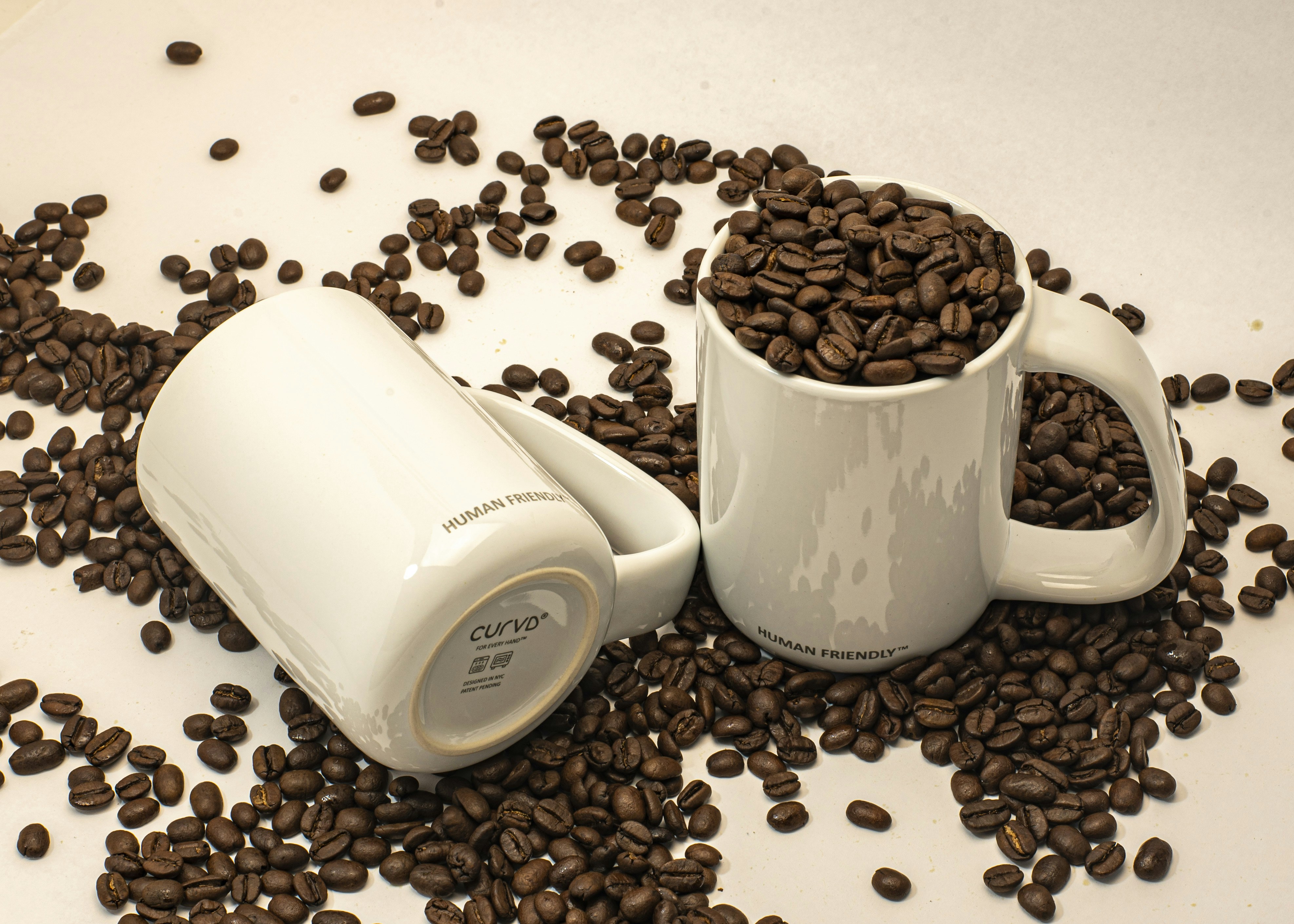 Two white mugs tipped over, spilling roasted coffee beans onto a white surface, creating an inviting scene for coffee lovers.