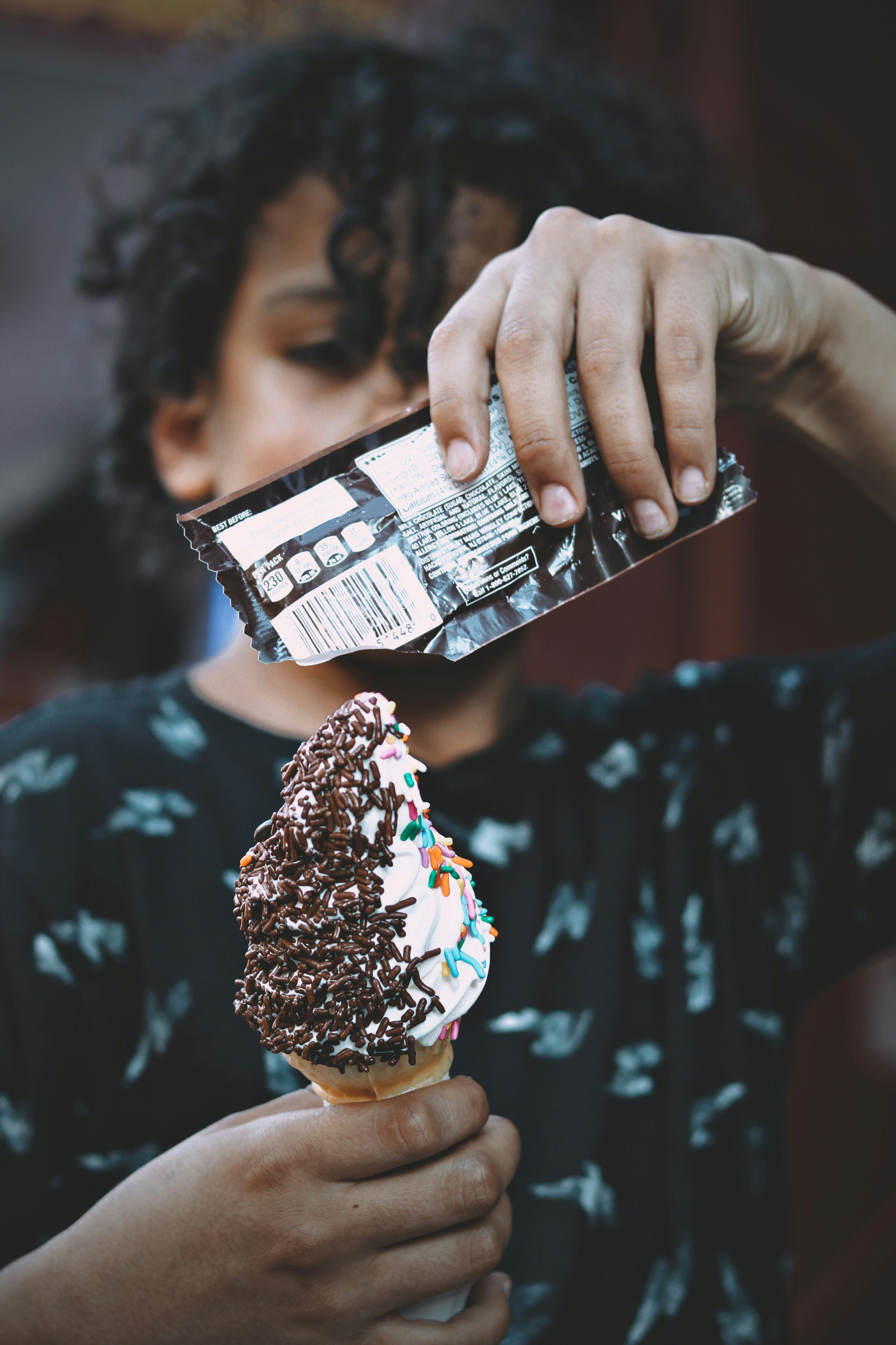 Child holding an ice cream cone topped with colorful sprinkles and chocolate, preparing to add more toppings from a packet.